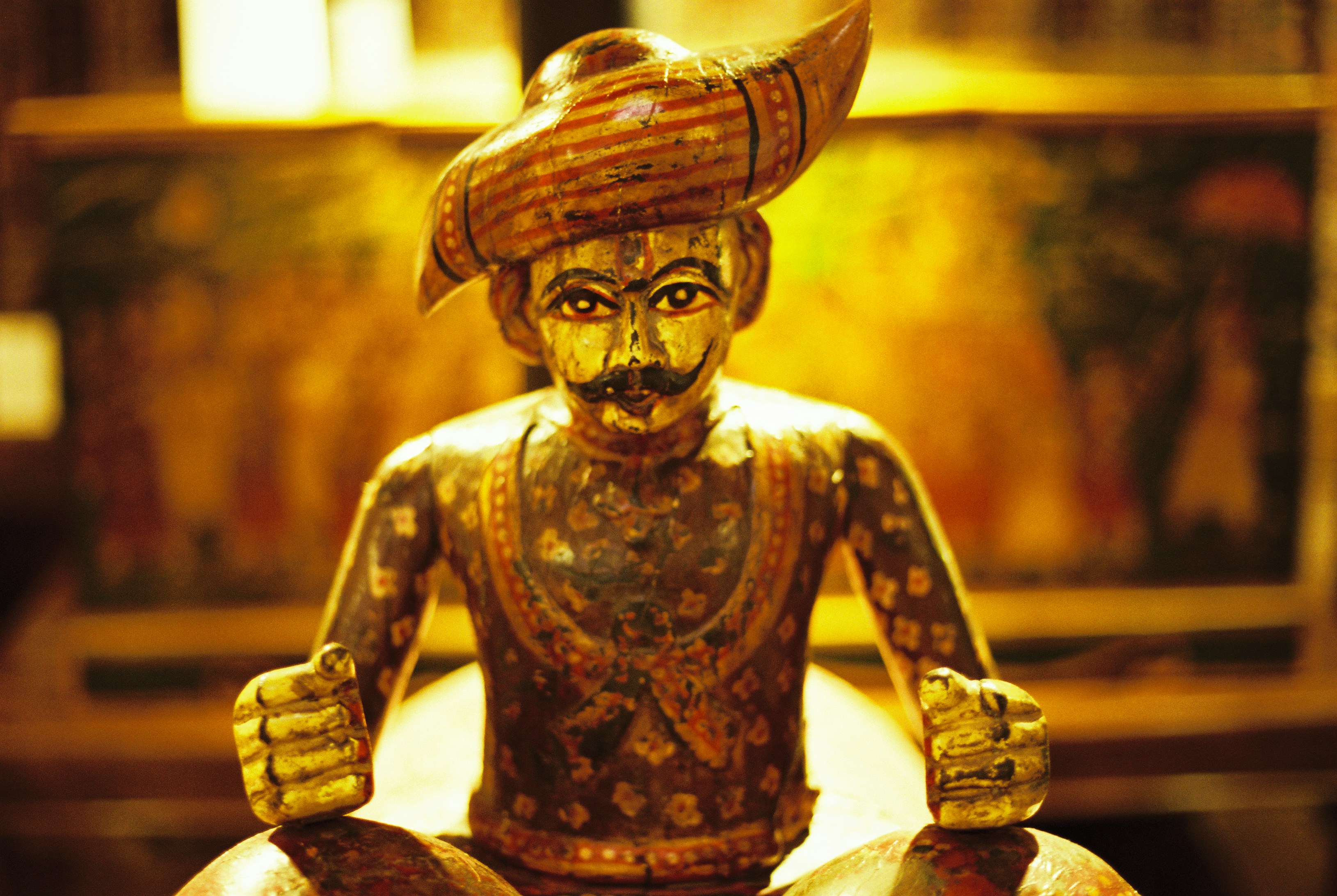 Carved wooden figure with a moustached face and ornate attire sits in a softly lit setting, captured as a compact portrait emphasizing its artful details and weathered texture.