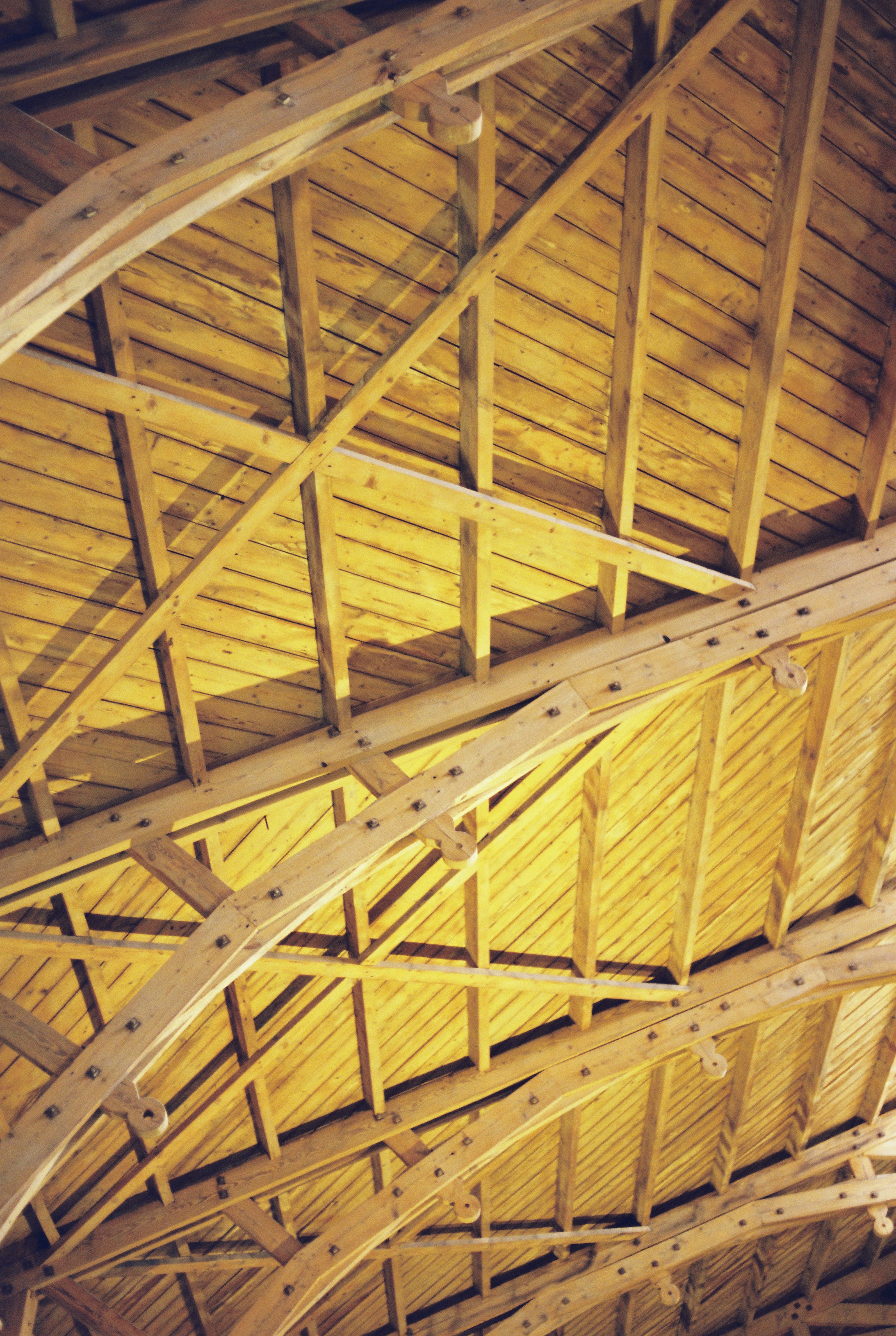 A close-up of sunlit wooden roof beams and trusses forming an intricate lattice with warm golden tones.