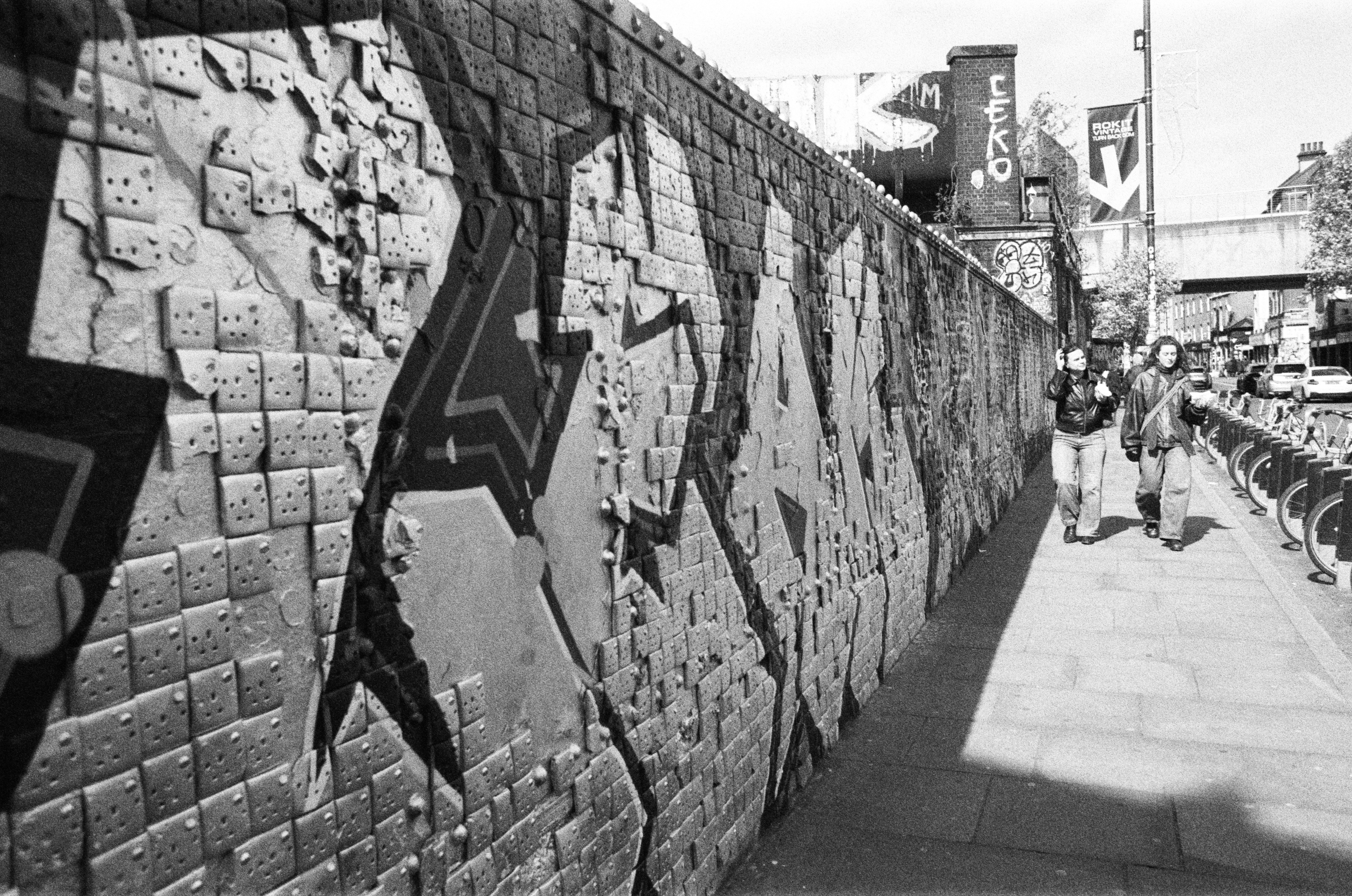 Graffiti-covered wall extends along a sunlit sidewalk with a few pedestrians in the distance; a city street scene captured in monochrome.