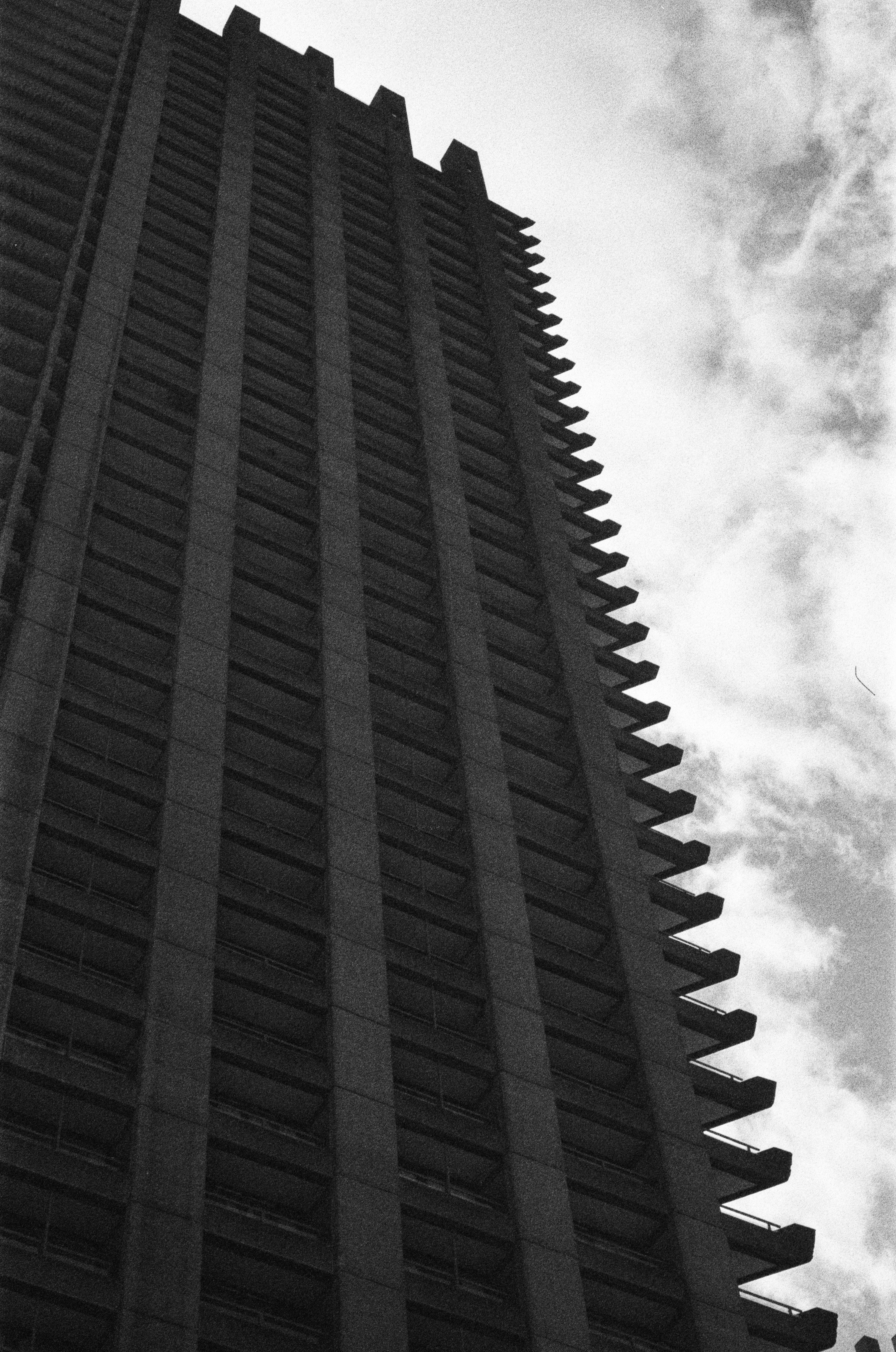 Tall, shadowed building facade rises along a diagonal, with repetitive balconies under a wispy sky. A stark, architectural close-up emphasizing form over color.
