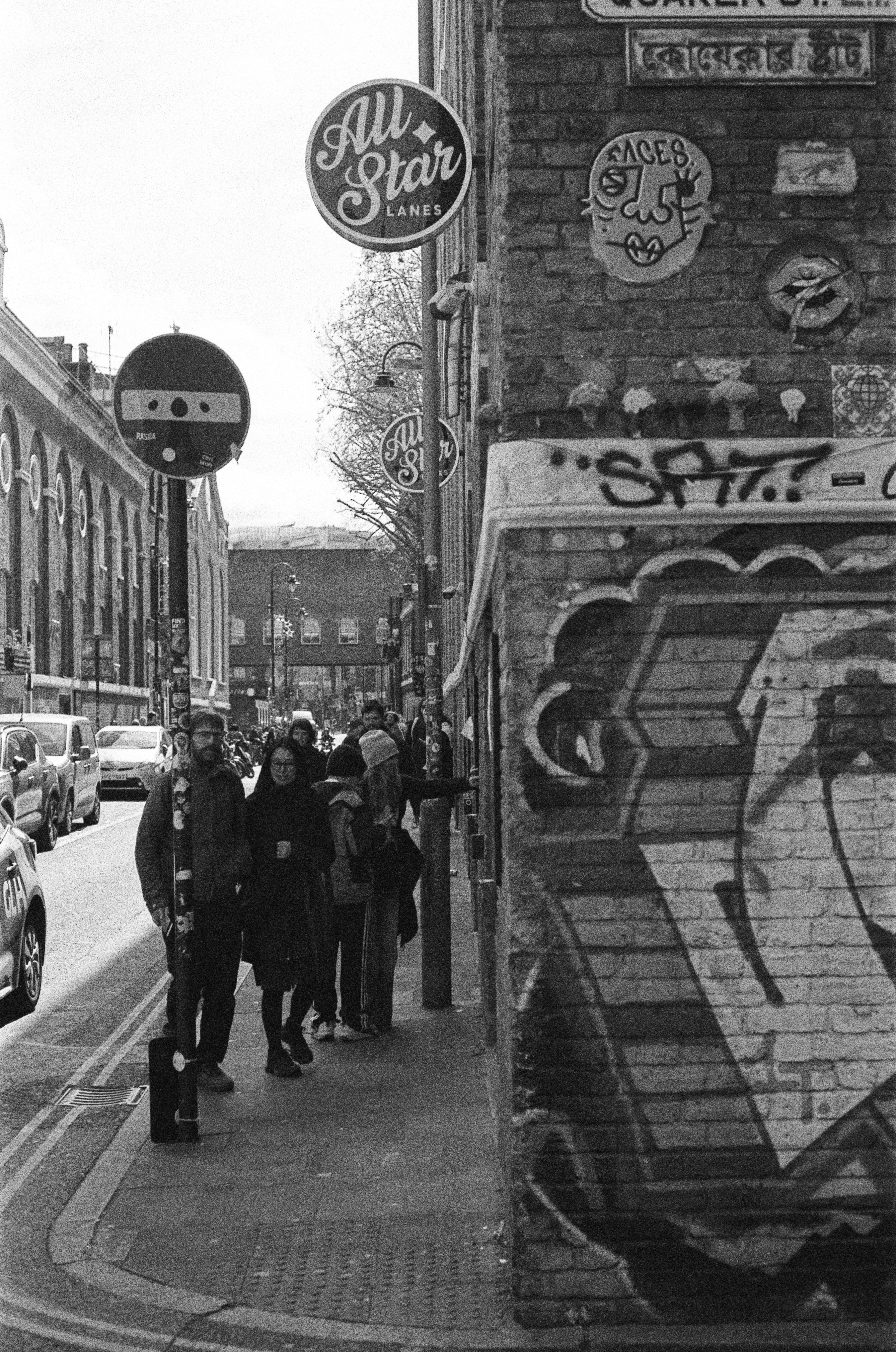 Urban street scene in black and white with a group of people waiting along a graffiti-covered wall and a vintage street facade.