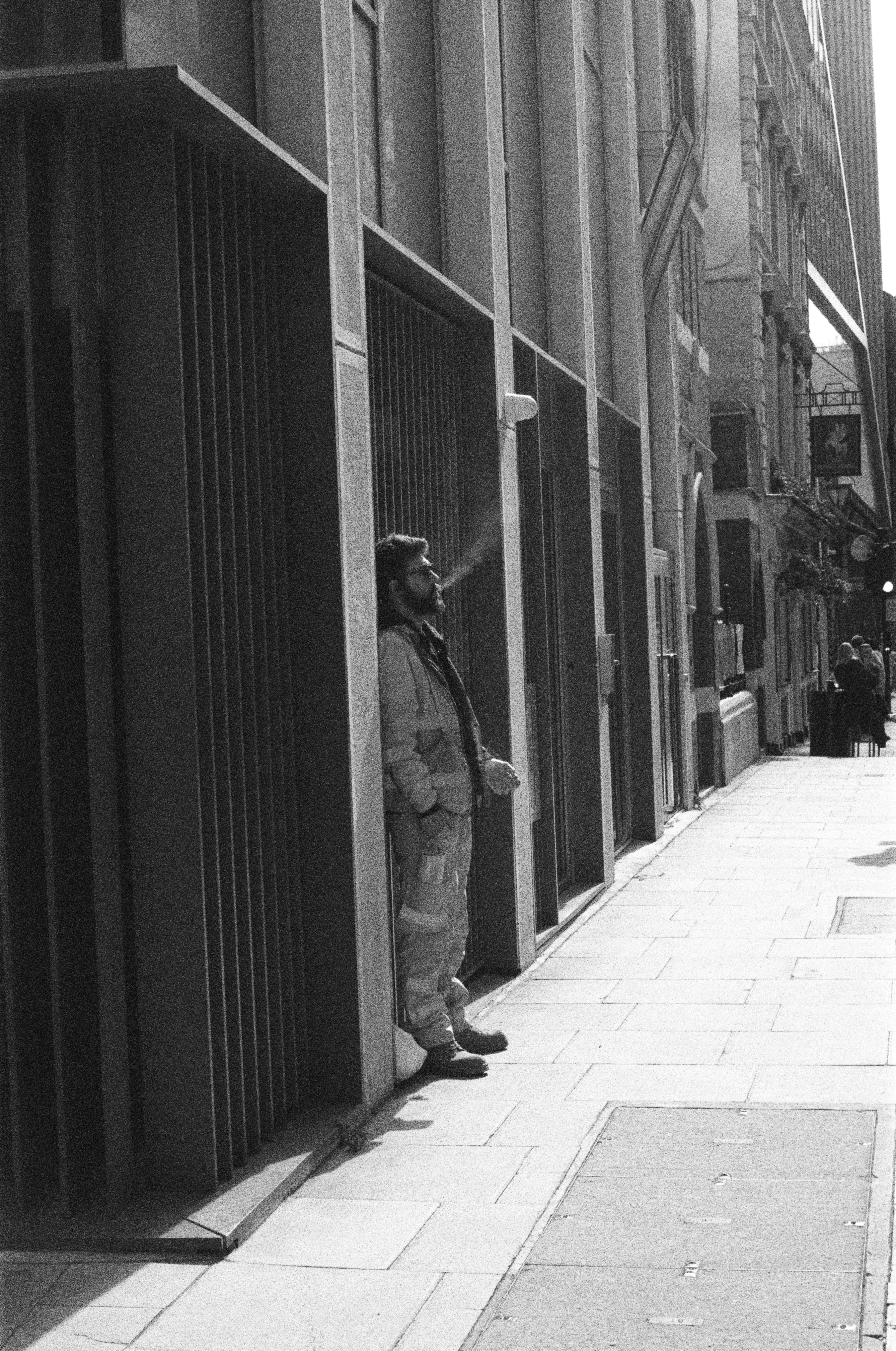 Man in a light jacket leans against a tall city façade as daylight pools along the sidewalk