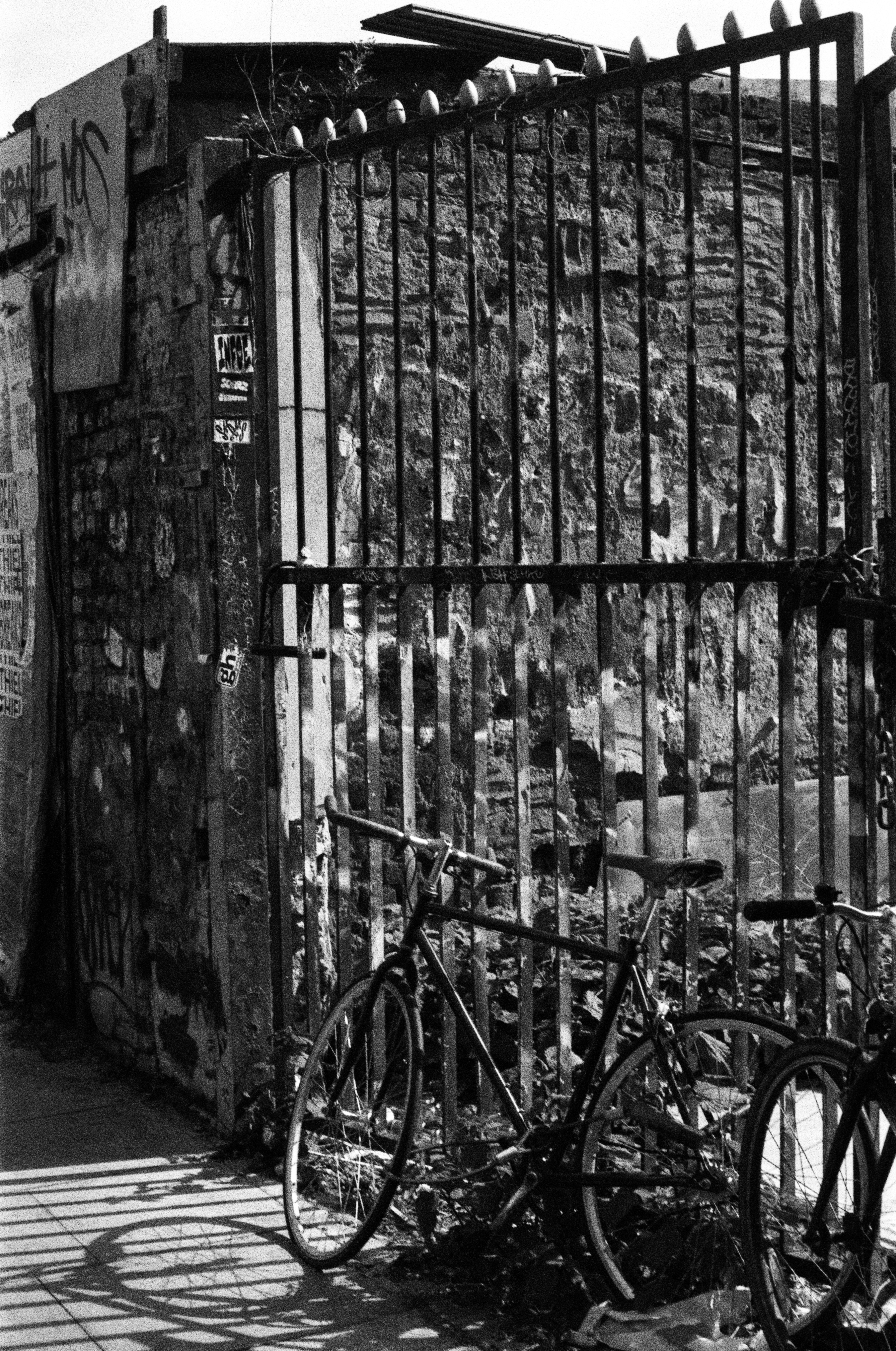 Dark alley scene featuring a tall metal gate with spikes and graffiti-covered walls, with bicycles leaning against the gate and a concrete, sunlit sidewalk in the foreground.