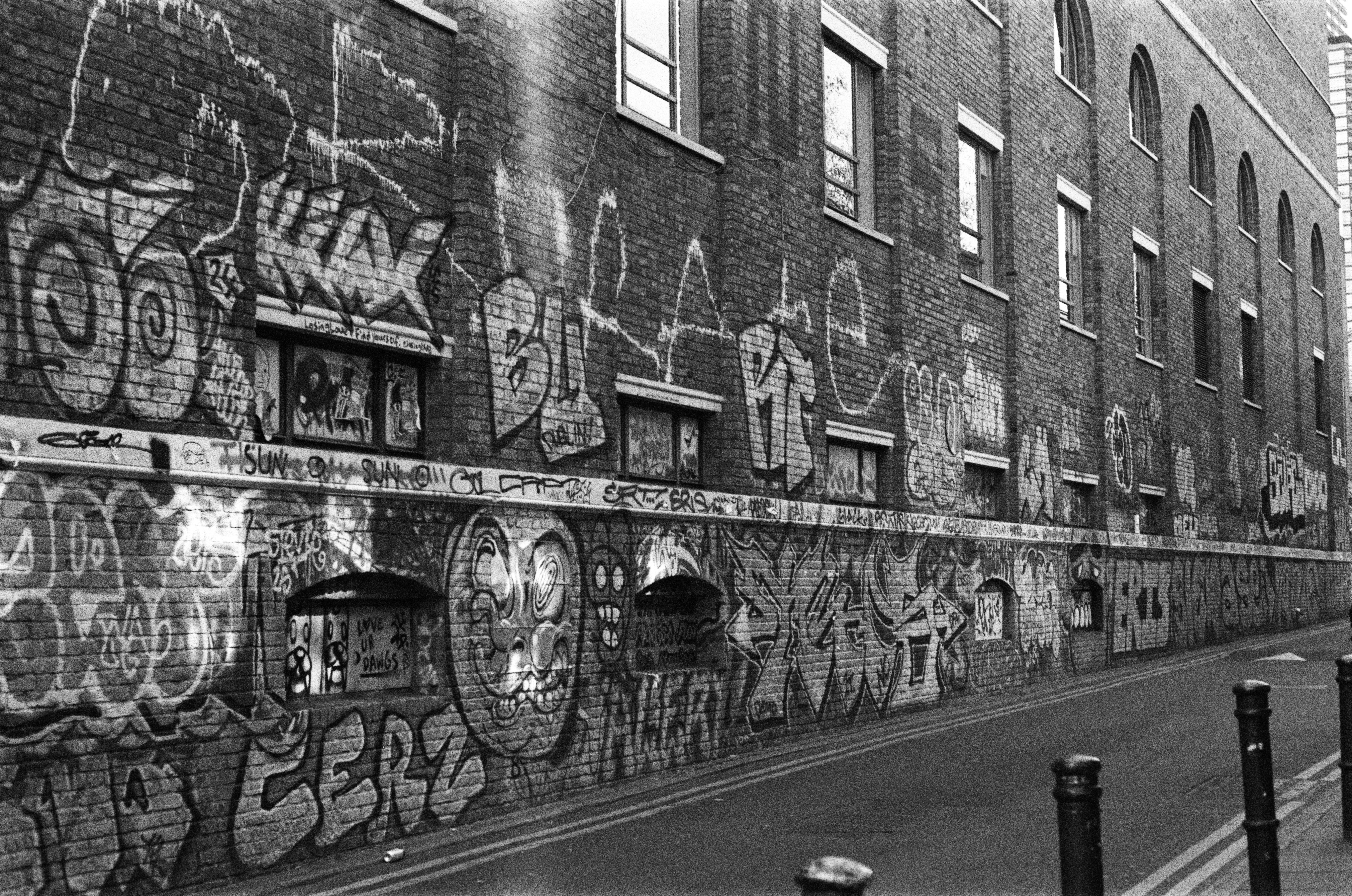 Monochrome alley with graffiti-covered brick wall and wet pavement, leading past bollards along a narrow street.