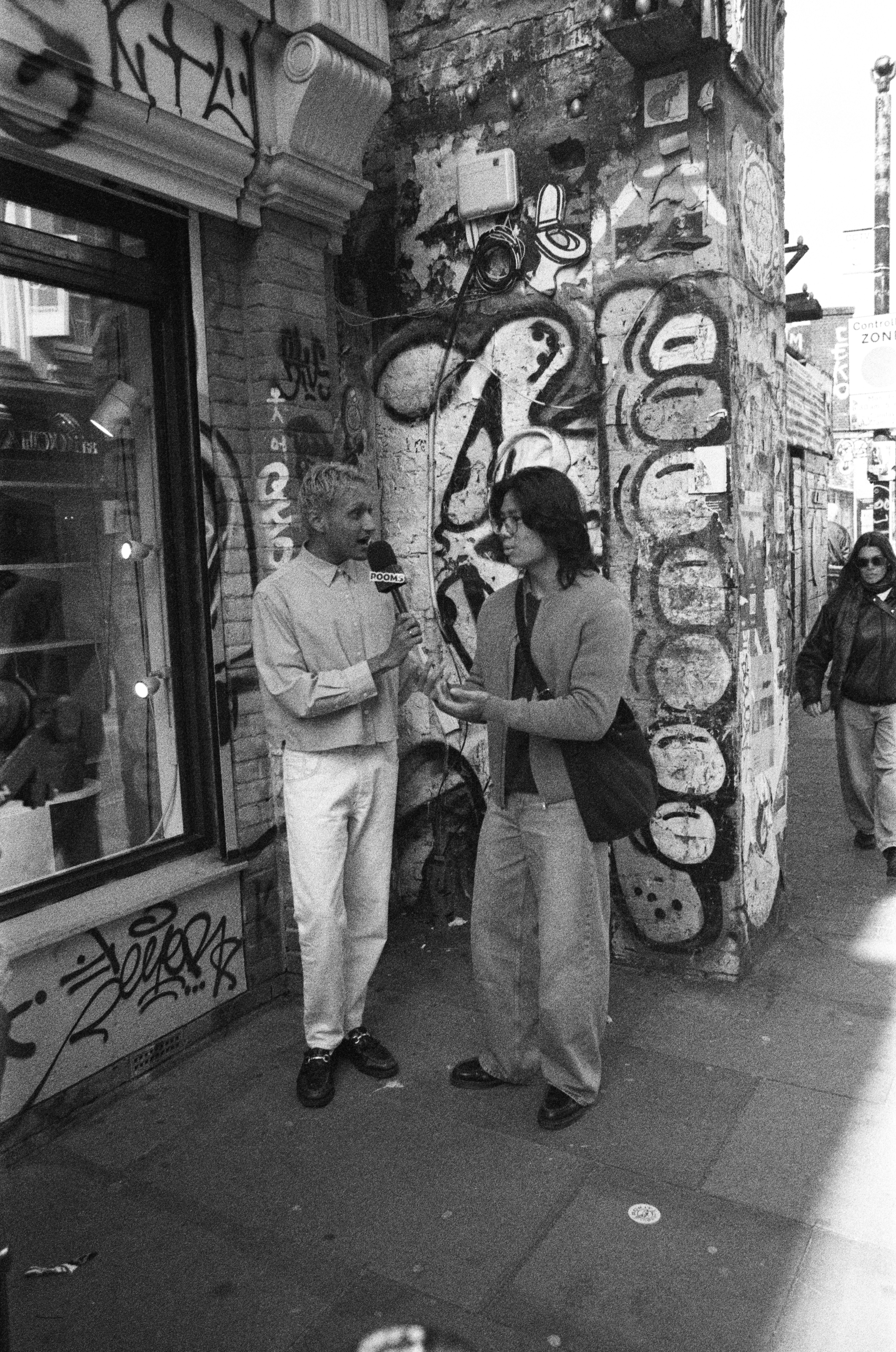 A man holding a microphone interviews a woman on a graffiti-covered sidewalk; a passerby walks by in the background near a storefront window.