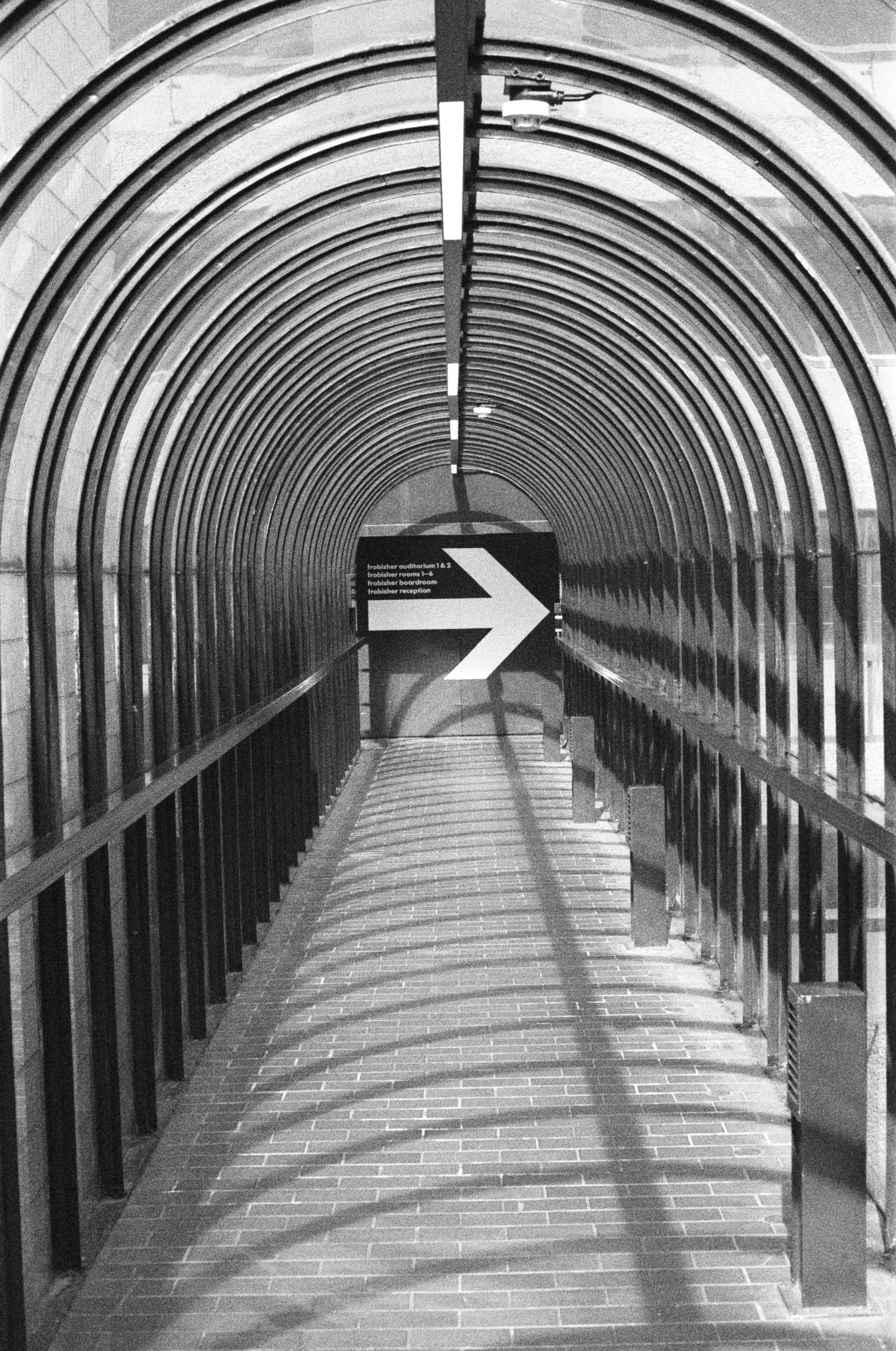 Monochrome tunnel with concentric arches leading to a bright arrow sign at the far end, casting geometric shadows on the brick floor.