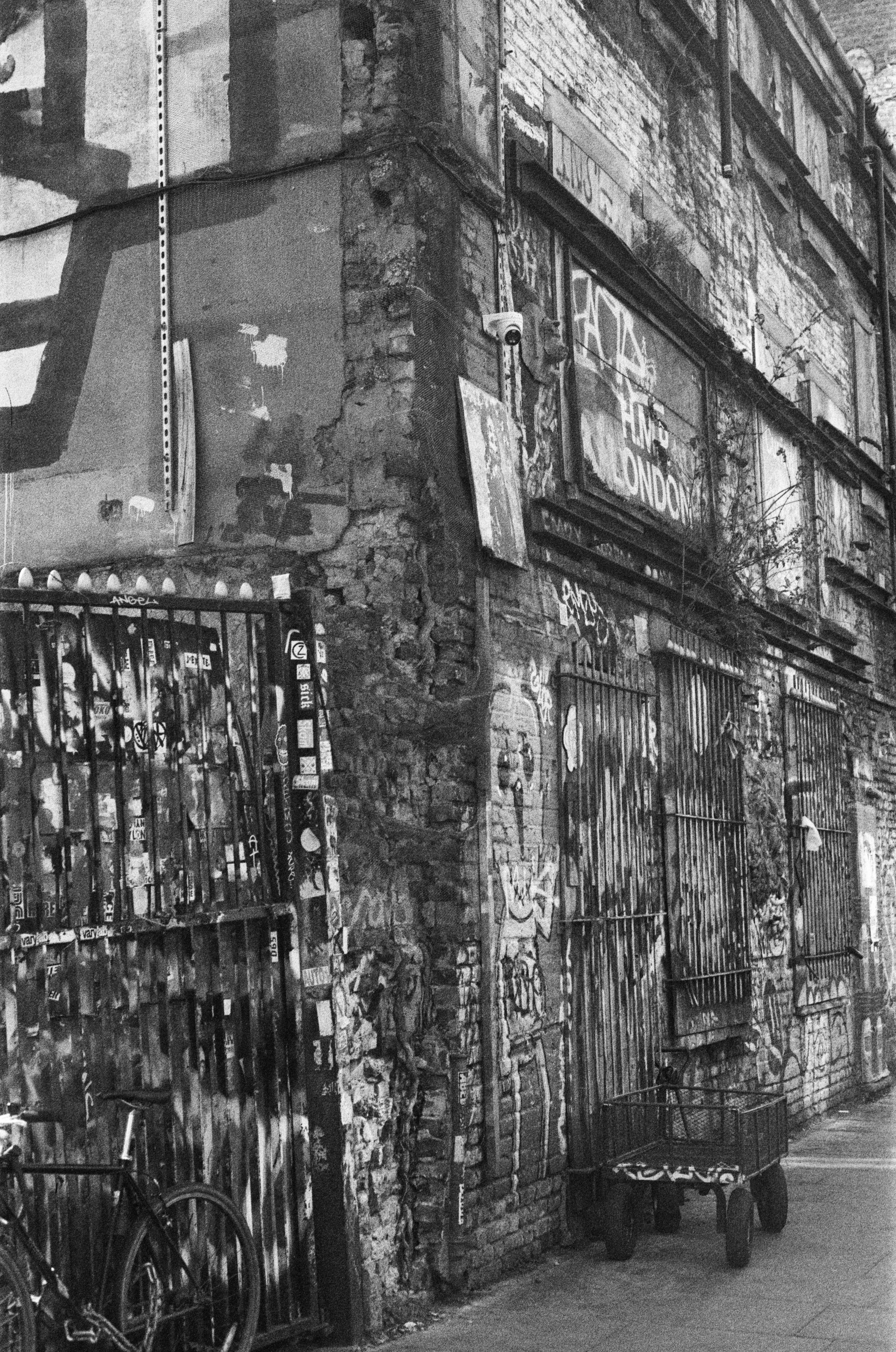 Monochrome urban alley with graffiti-covered brick walls, a rusted cart and a bicycle against a gated doorway, conveying grit and texture.