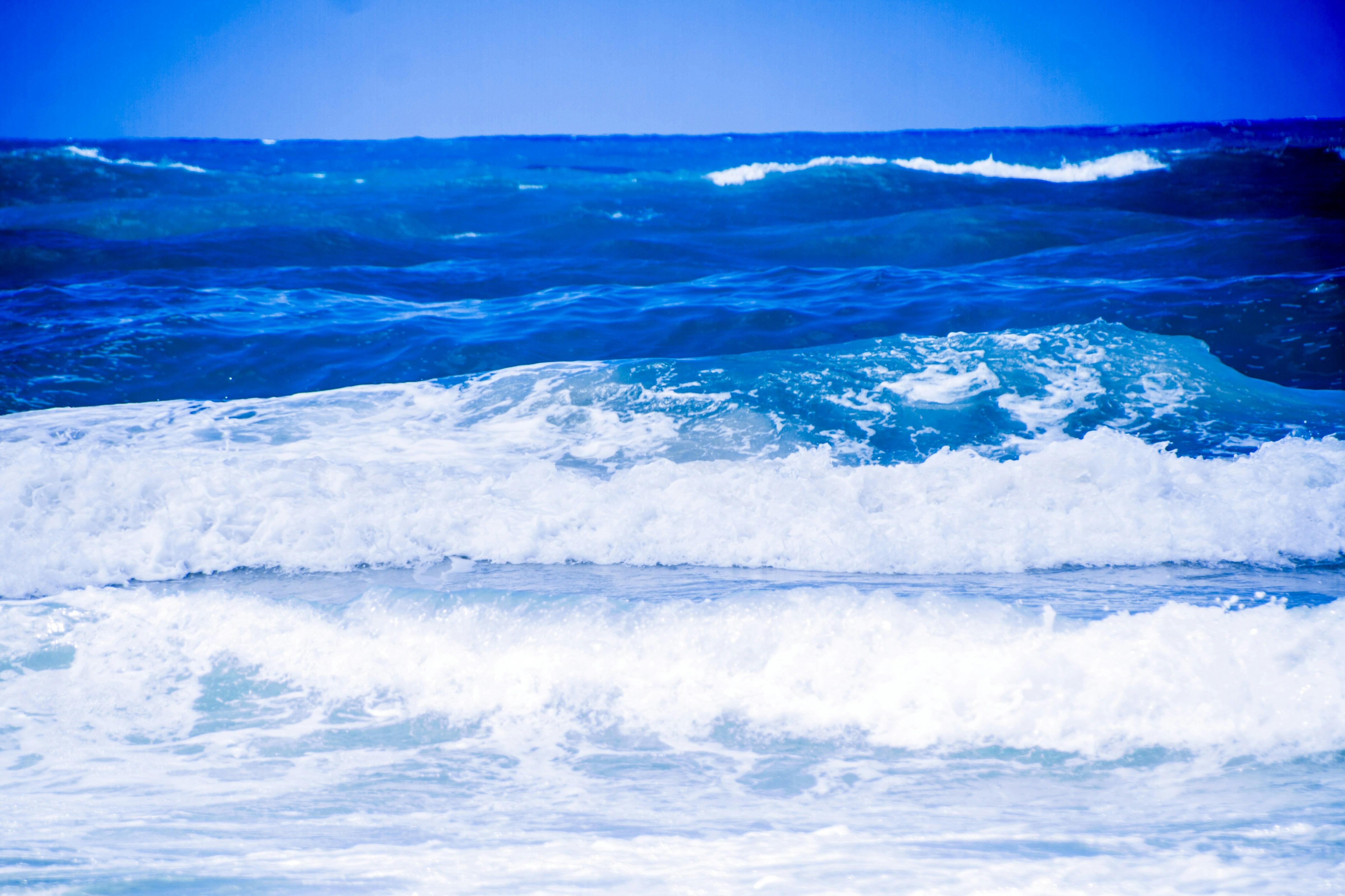 Blue ocean waves crest and break along a sunlit shoreline, captured with emphasis on the foamy white spray and varying turquoise hues. A minimalist seascape with a focus on motion and texture.