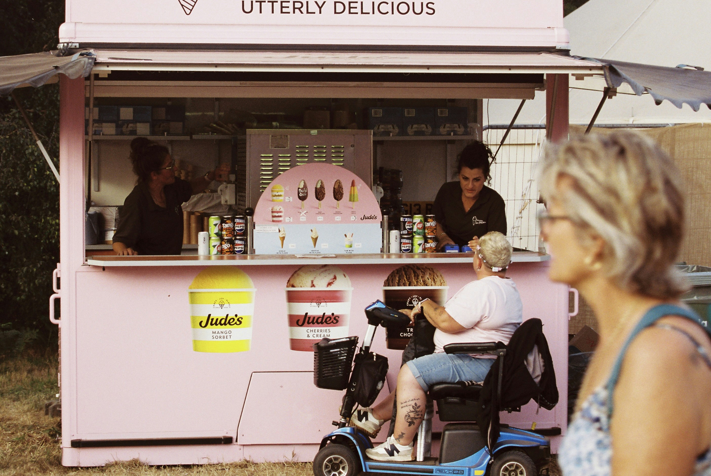 A pink food stand with two attendants operates at a fair, while a person on a blue mobility scooter is served; a blurred foreground figure passes by on the right. This is a candid street scene with emphasis on the stand and its customers.