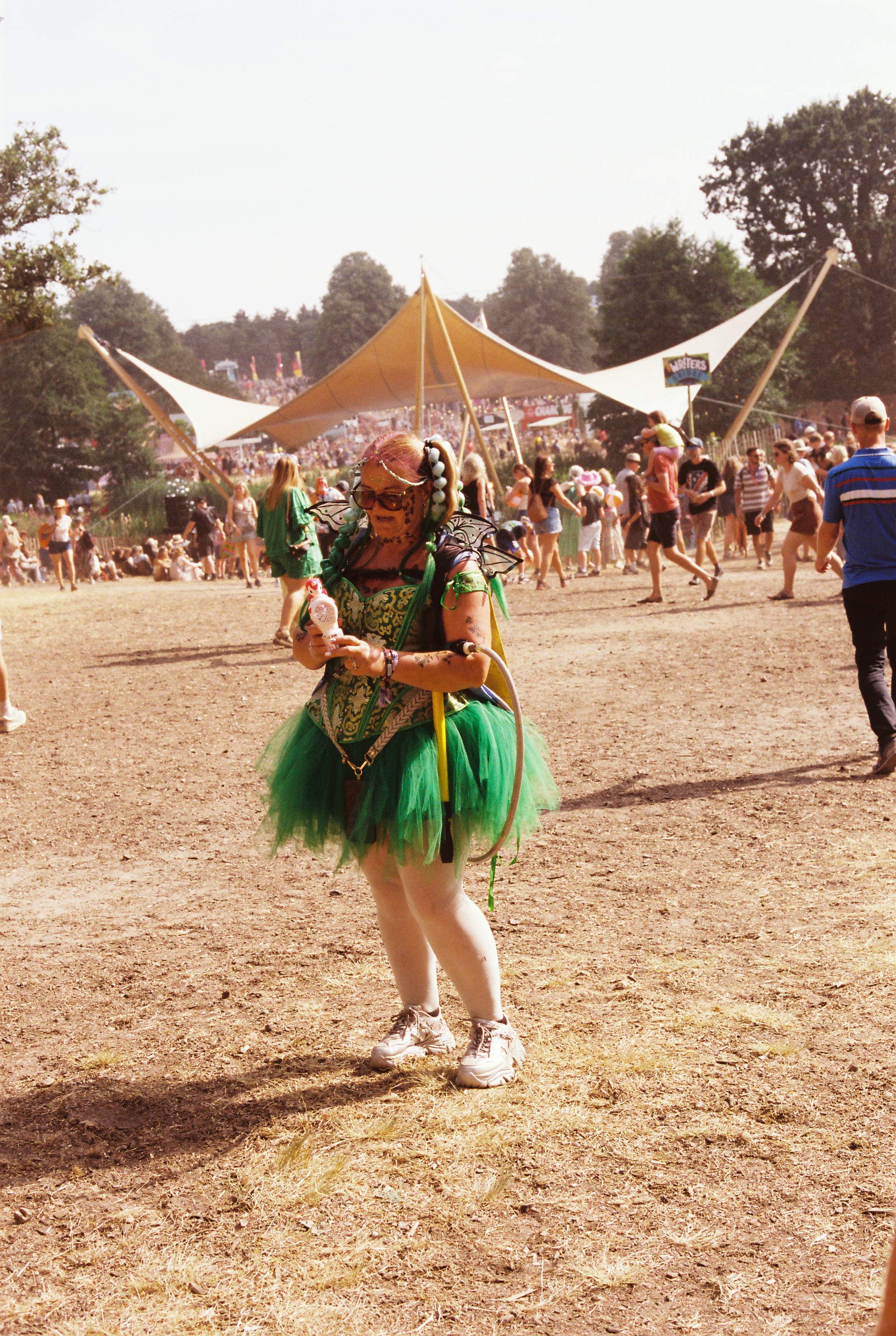 A costumed performer in a green tutu stands center-stage at a sunny outdoor festival, backgrounded by a crowd and festival tents.