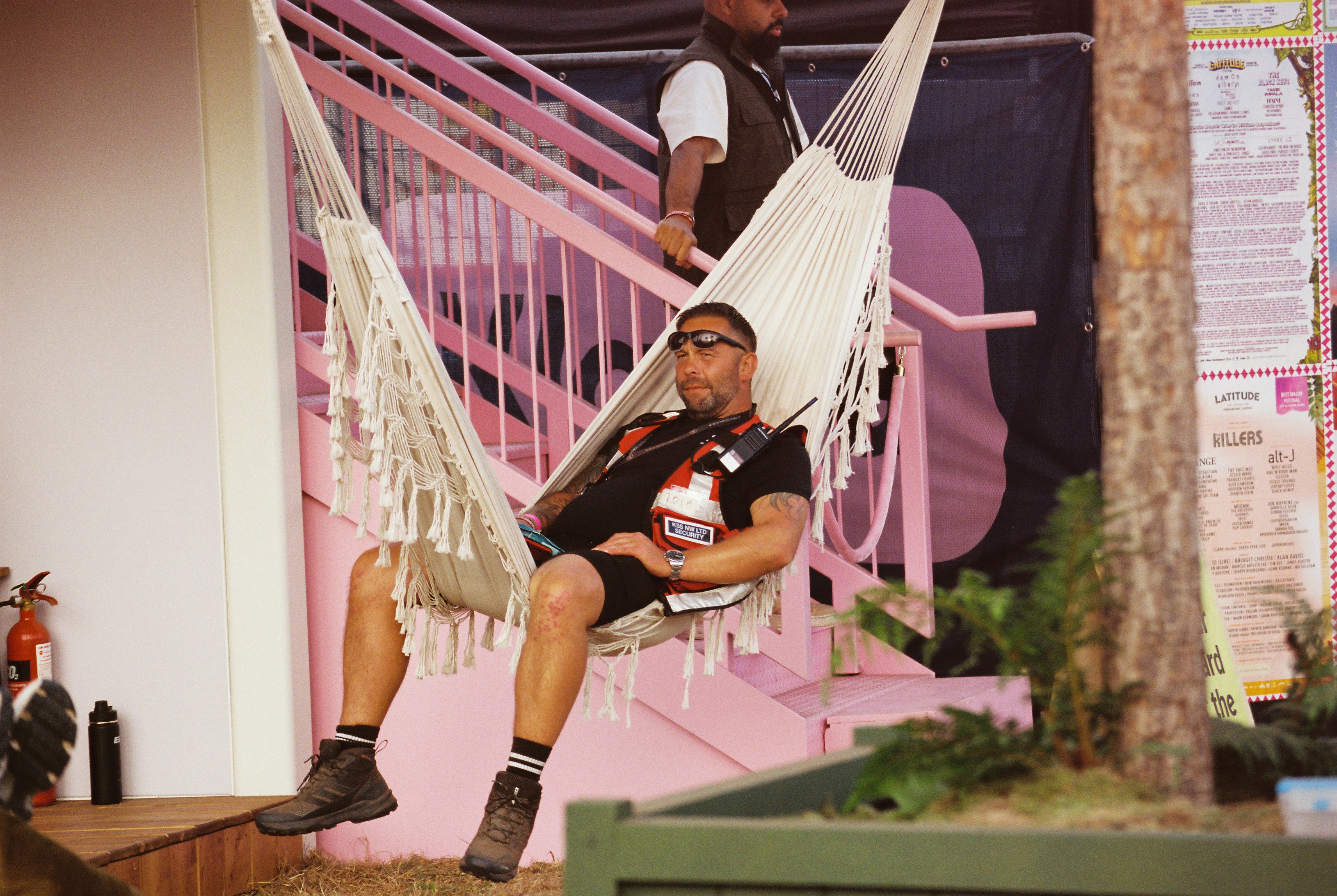 Man resting in a white hammock with pink accents, wearing sunglasses and a dark shirt; background shows a pink staircase and a poster-adorned wall, suggesting a casual outdoor venue.