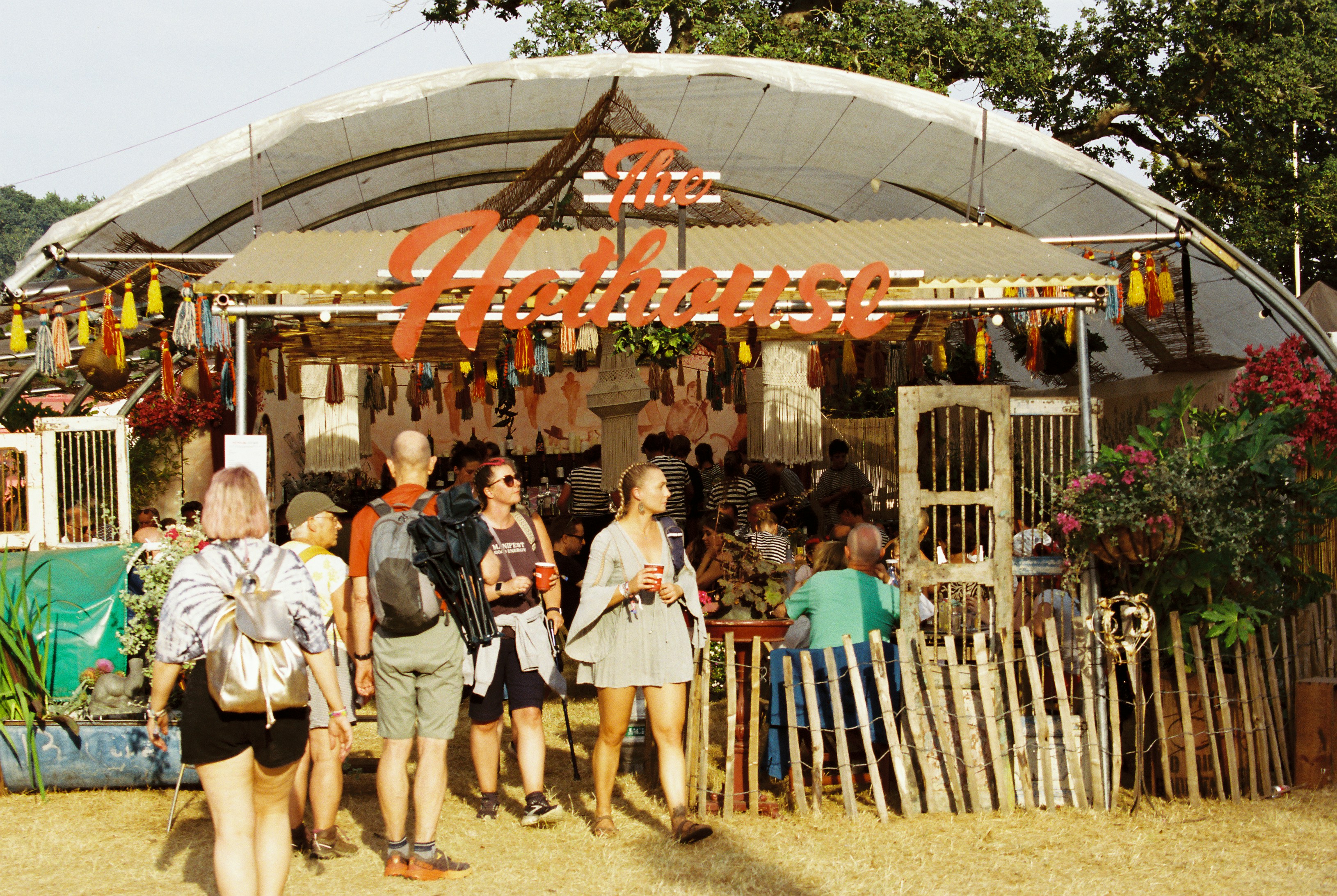 A festive outdoor bar or cafe scene under a white canopy with a bright orange sign; people mingle near rustic fencing and plants, creating a lively social moment.