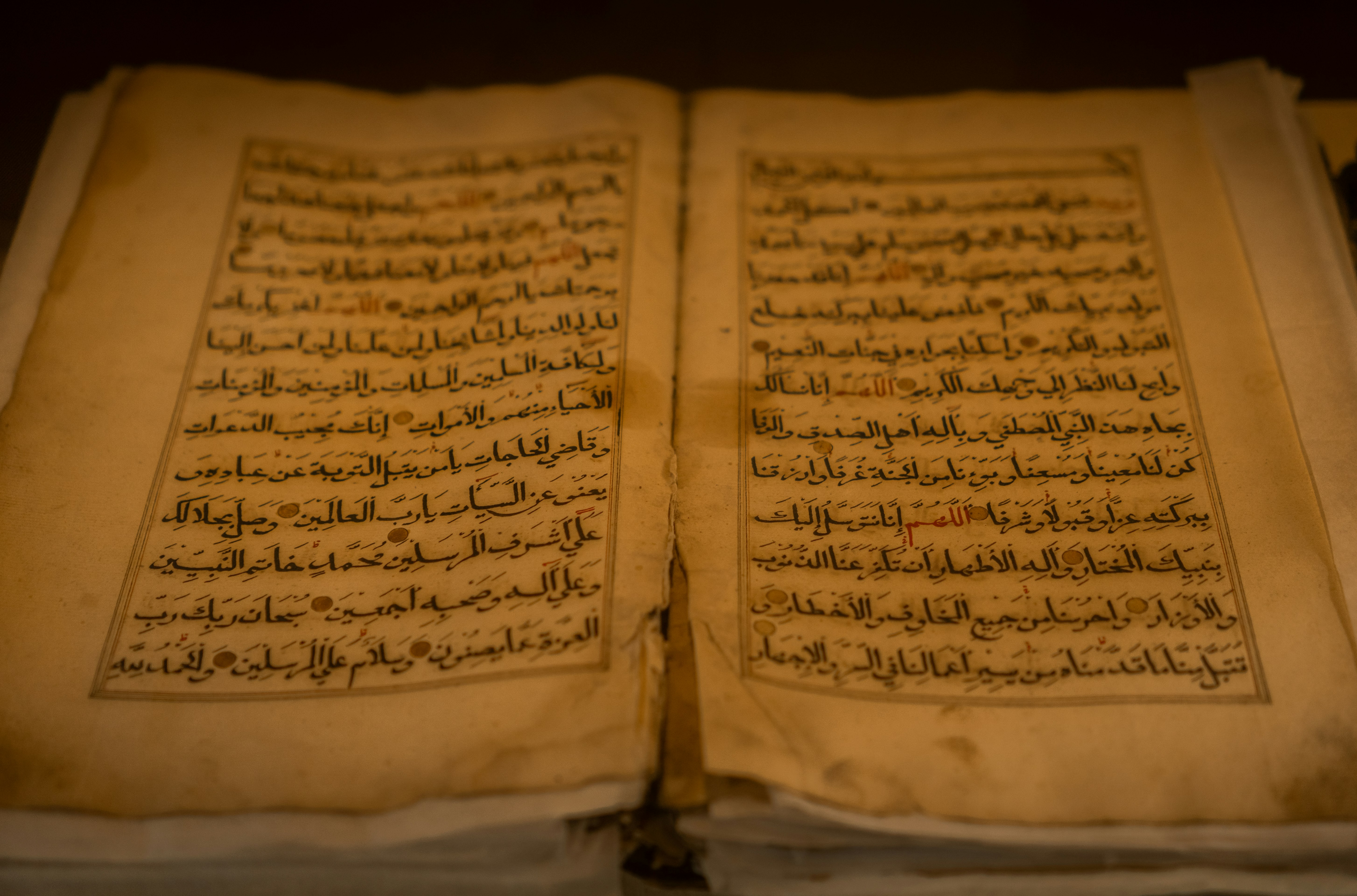 Two open aged manuscript pages with Arabic script, leather binding at the bottom, soft warm light; appears to be a historical or religious text with visible wear and foxing.