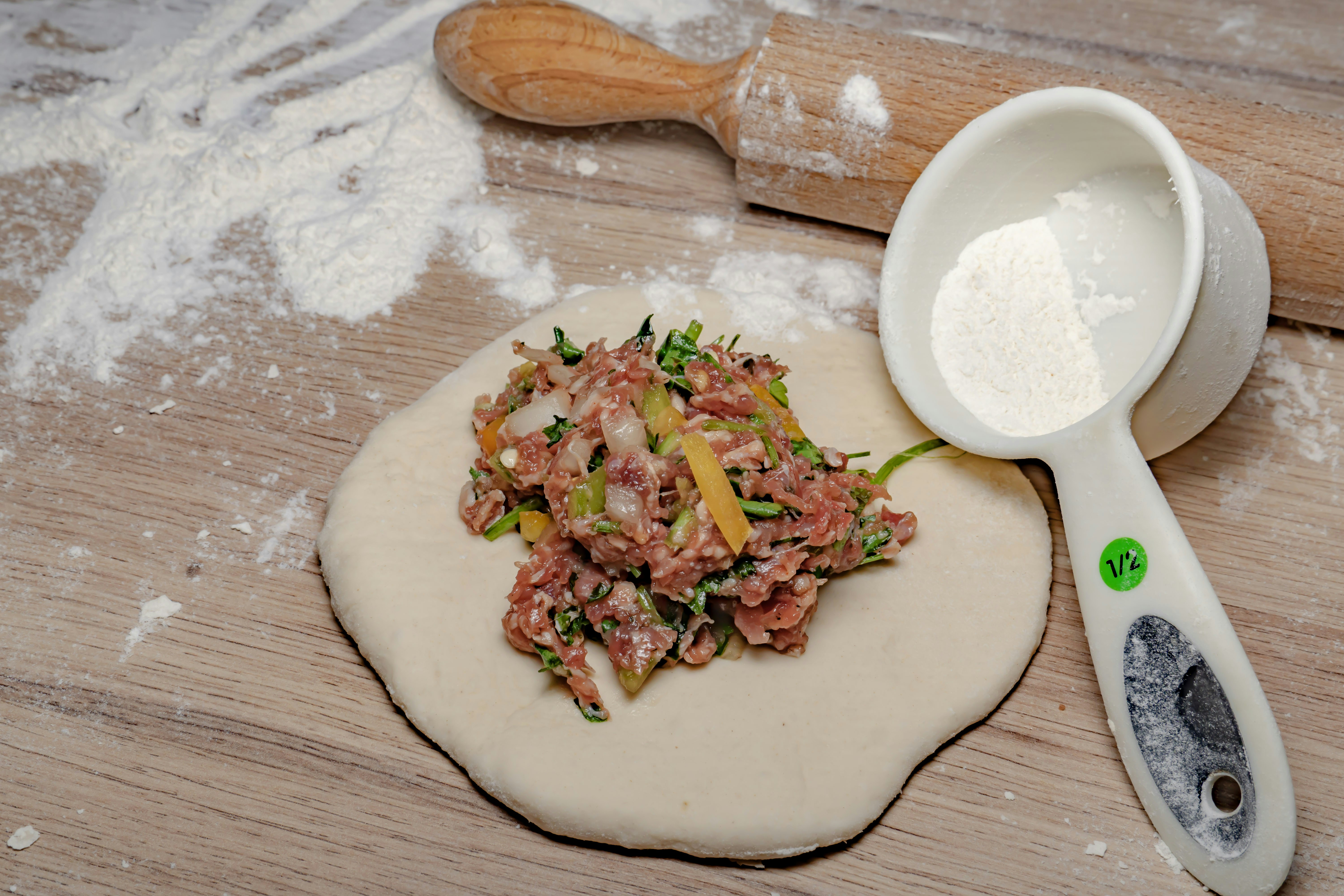 Dough with a minced filling placed on a wooden work surface, surrounded by flour and a measuring scoop with a mound of flour. The image captures the preparation stage of handmade pastry.
