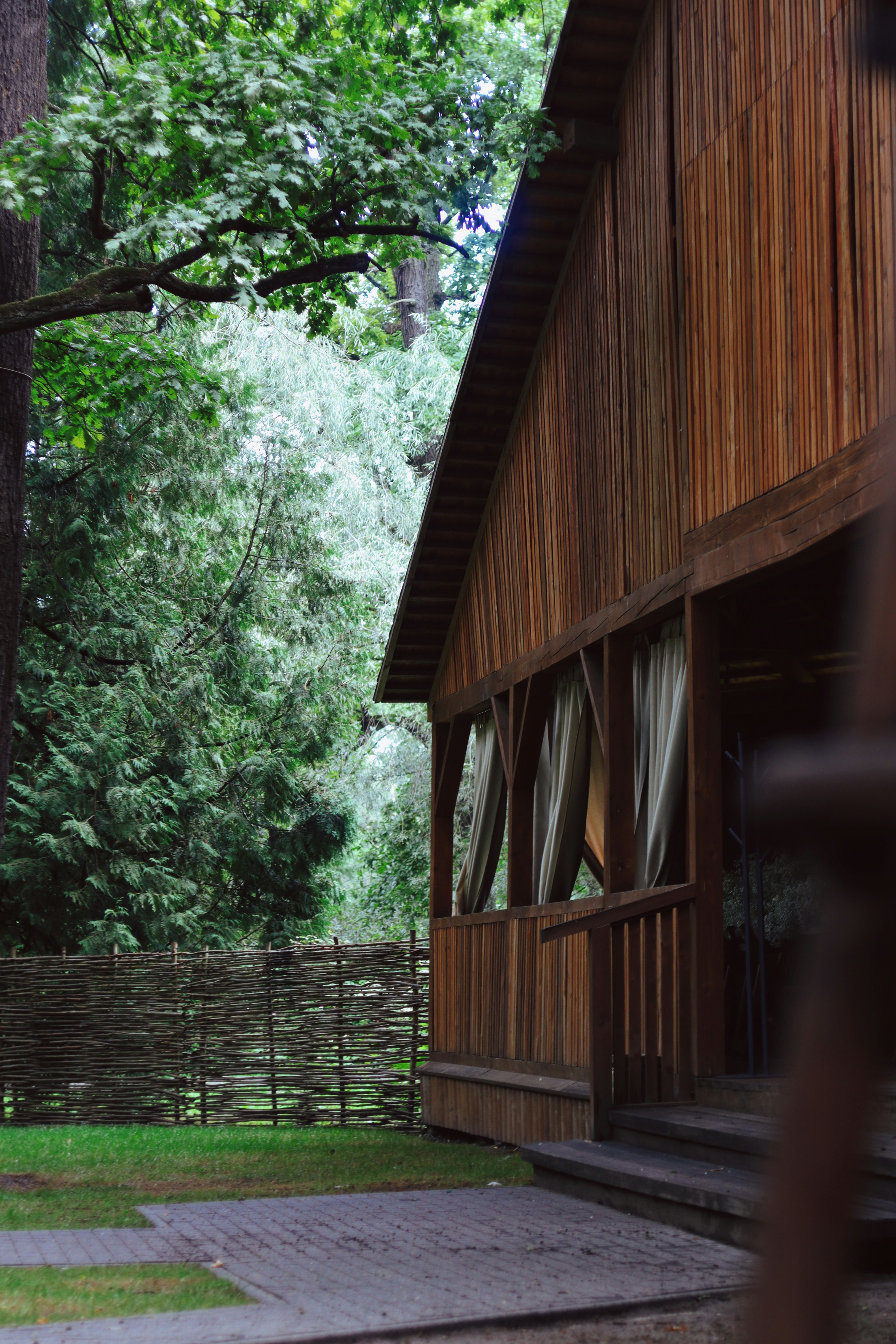 A rustic wooden barn-style pavilion with a vertical slat facade and open windows with curtains, nestled next to a traditional wicker fence and lush green trees in a countryside park.