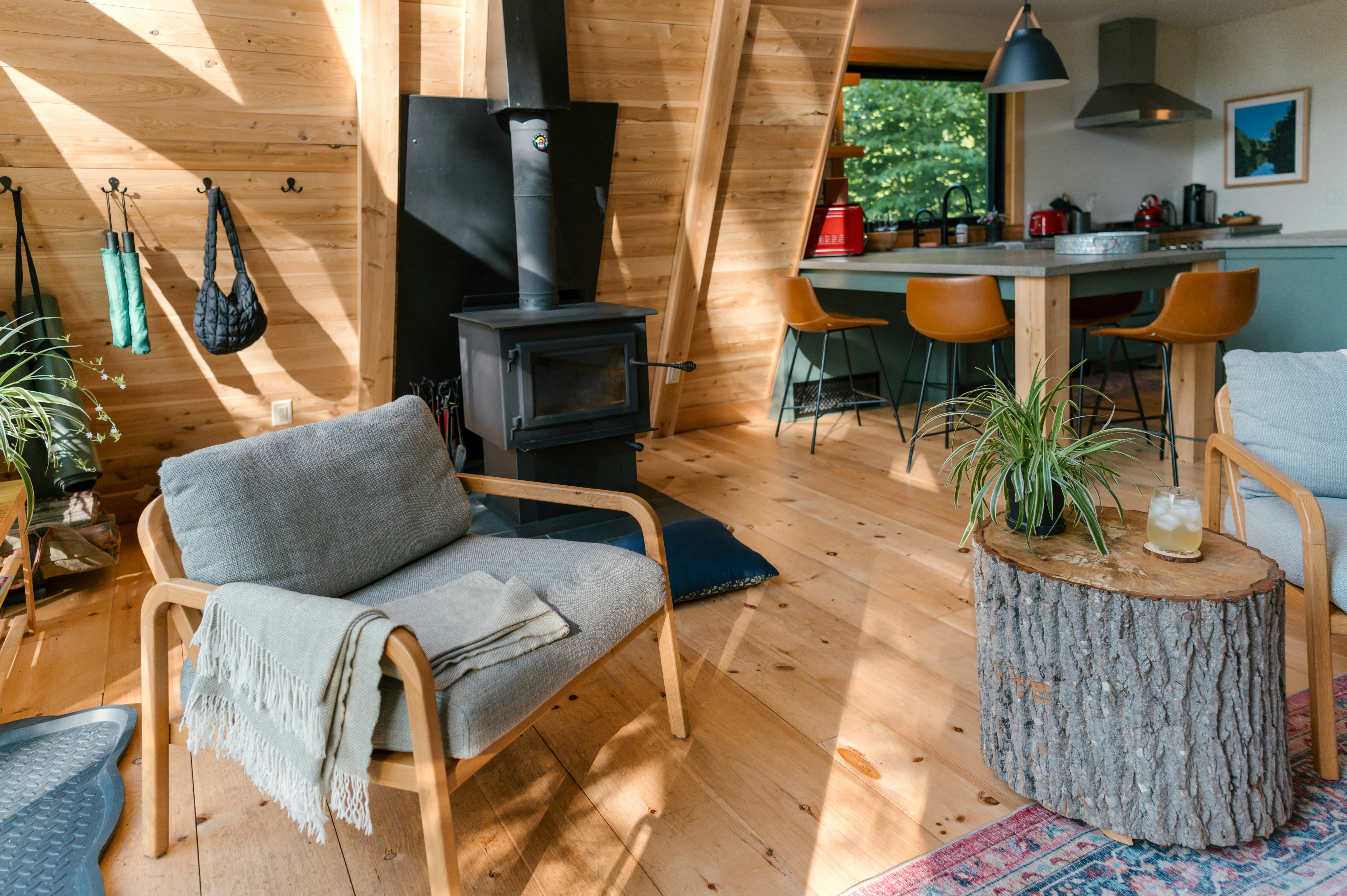 Warm, sunlit living space with a wood stove, architectural timber walls, and a mid-century chair setup surrounding a reclaimed log coffee table.