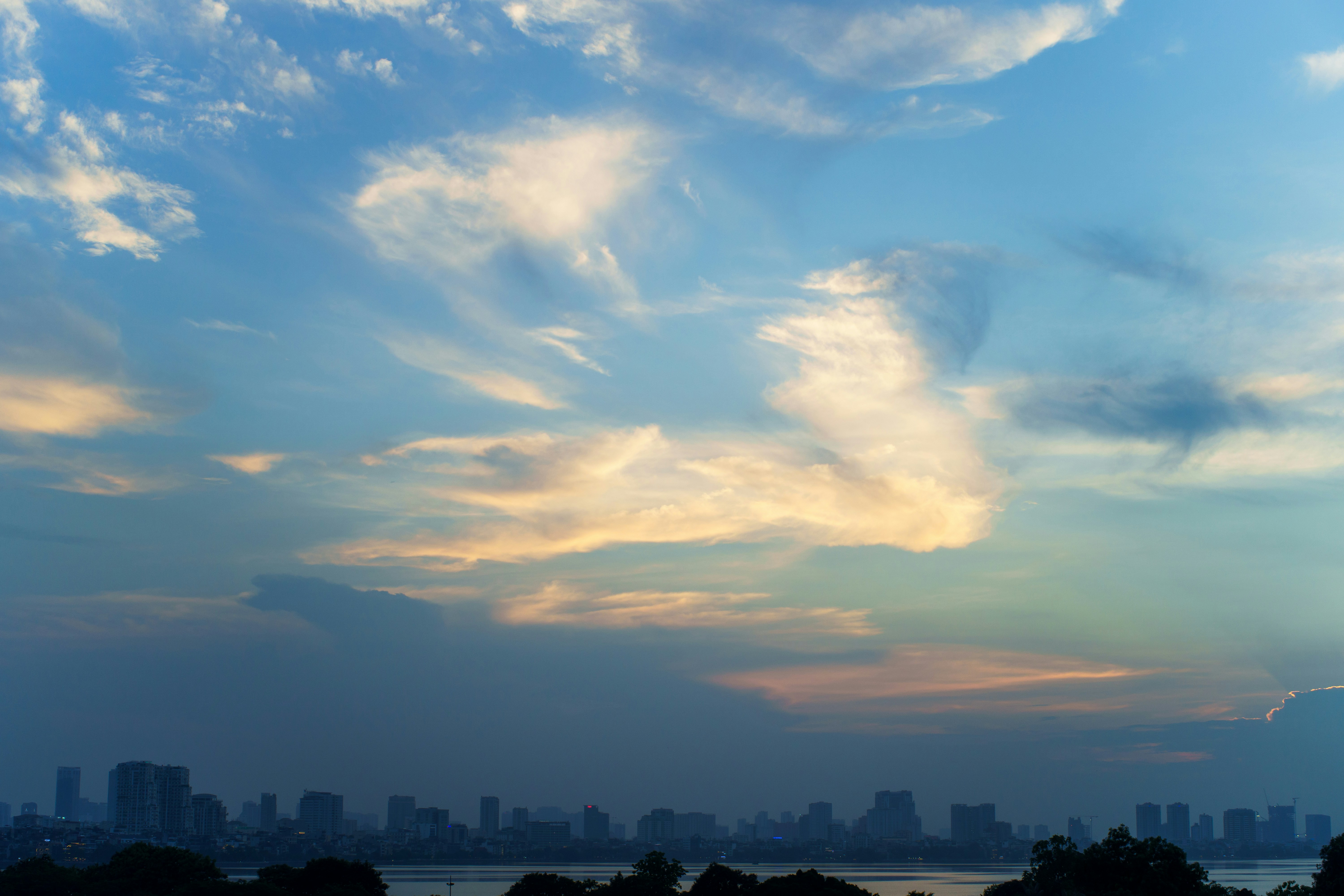 Twilight sky over a calm waterfront with a distant urban silhouette and low-lying vegetation in the foreground. The scene emphasizes serene color gradients and a soft horizon line.