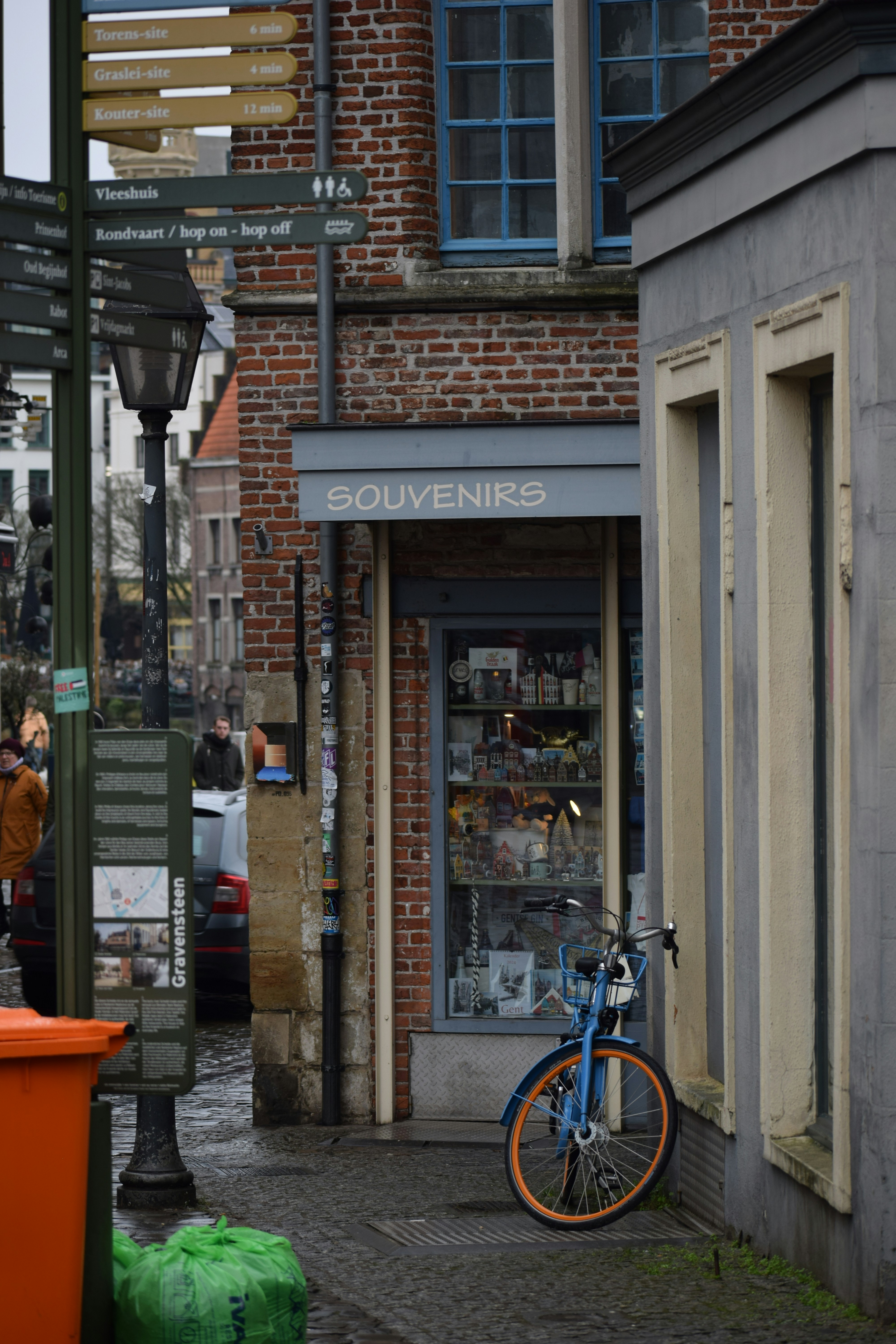 A brick-fronted souvenir shop sits along a narrow cobbled street, with a blue bicycle leaning against the wall beside large display windows. Street signs and a bright orange bin anchor the left, while a stone building frames the right.