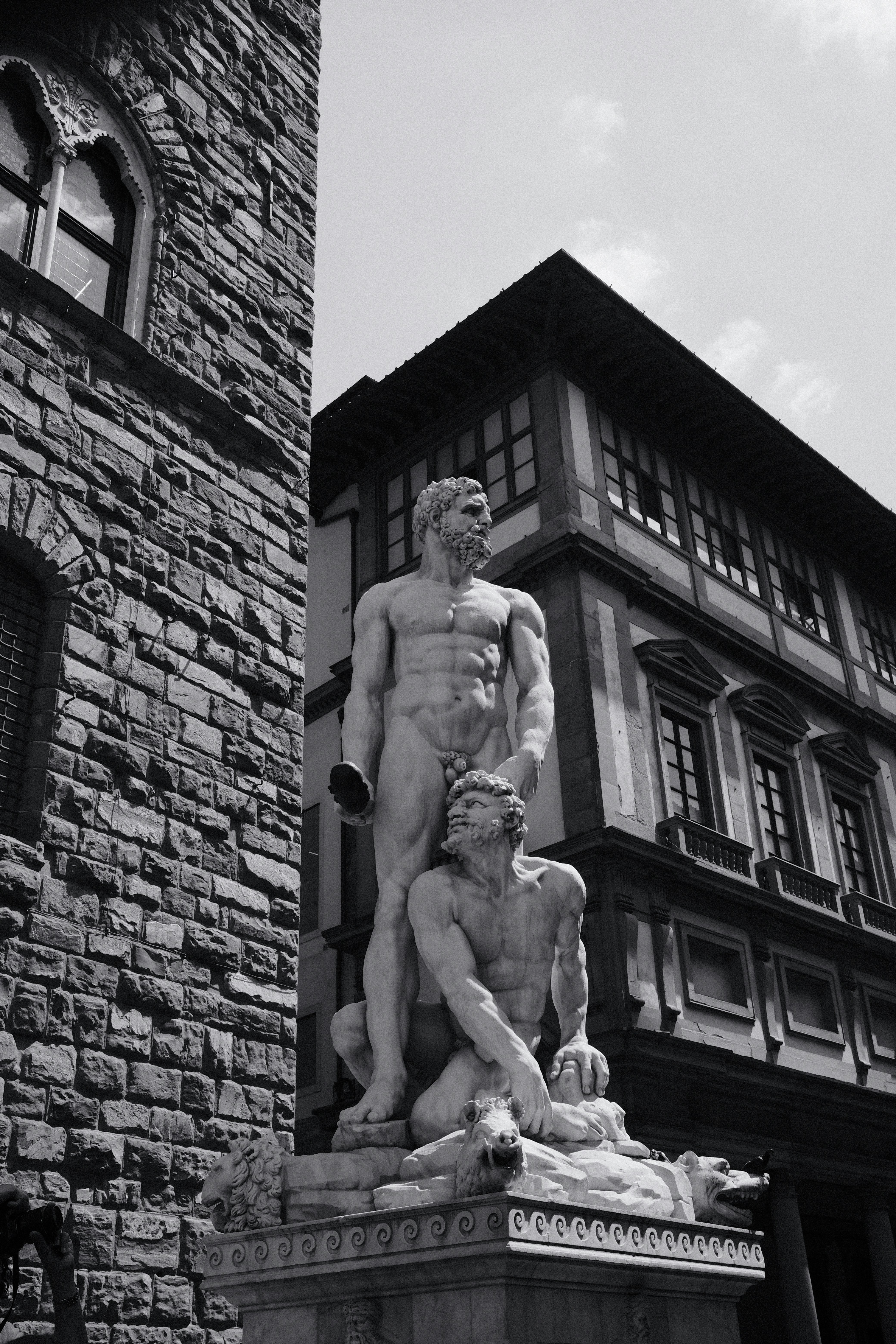 Monumental marble statues stand centered against historic stone and timber architecture in grayscale, emphasizing classical sculpture within an urban facade.
