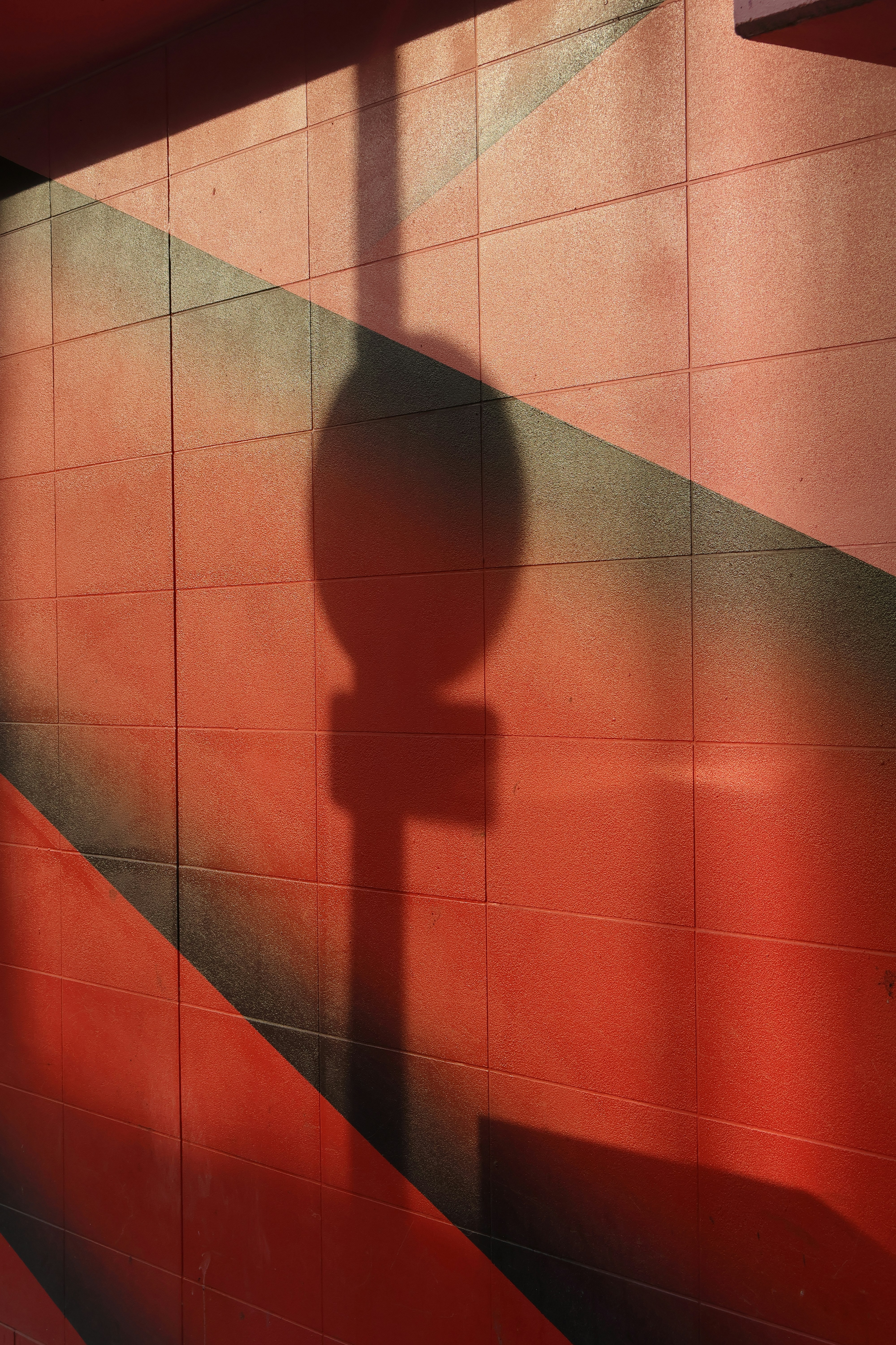 A large circular shadow with a vertical post sits on a red-tiled wall, intersected by diagonal shapes and soft color gradients. This image emphasizes geometric shadows and architectural texture.