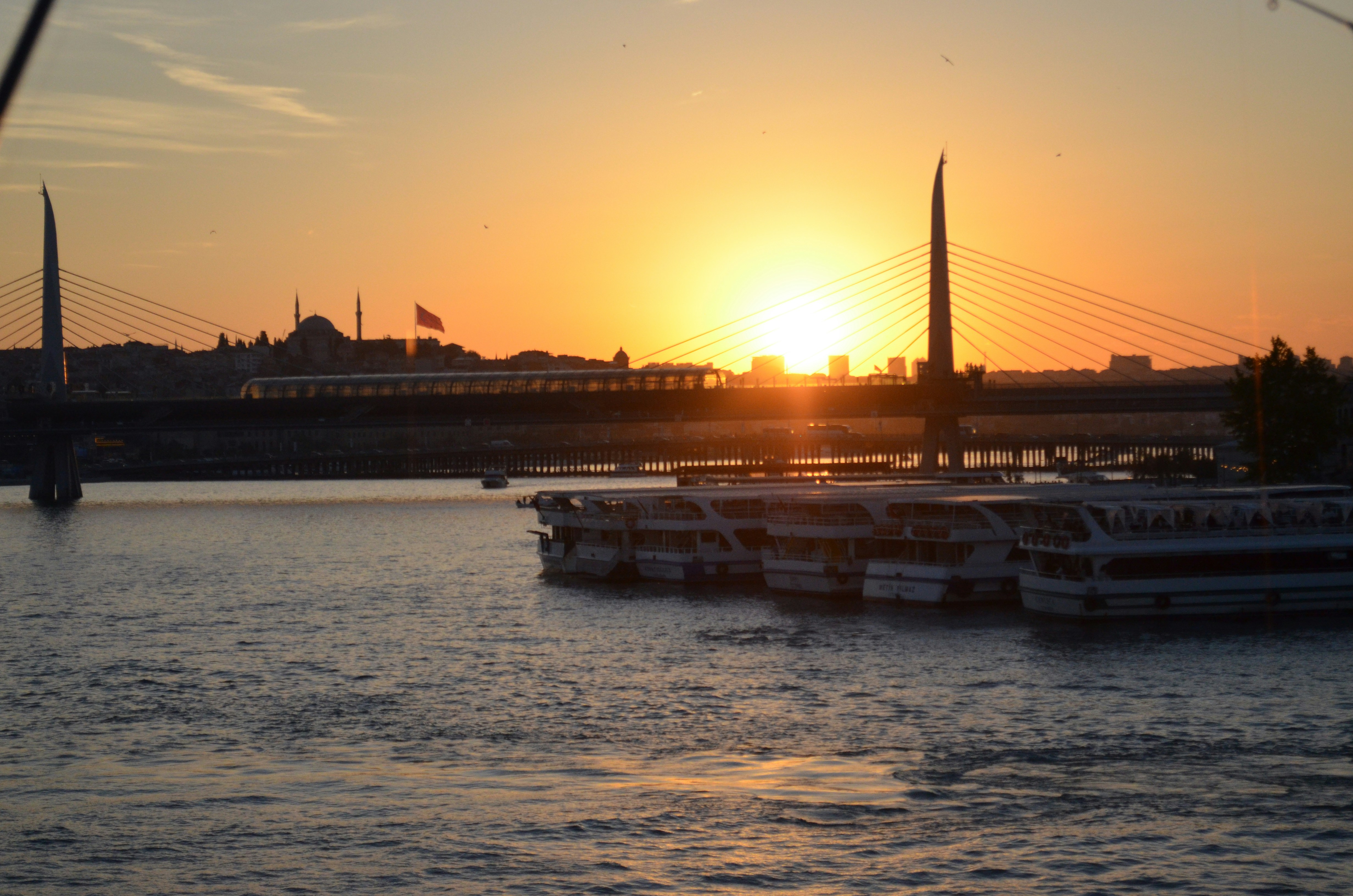 Sunset over a river with a bridge silhouette and boats docked along the foreground; a calm scene captured near dusk with warm tones. This is a photographic frame of a real scene.