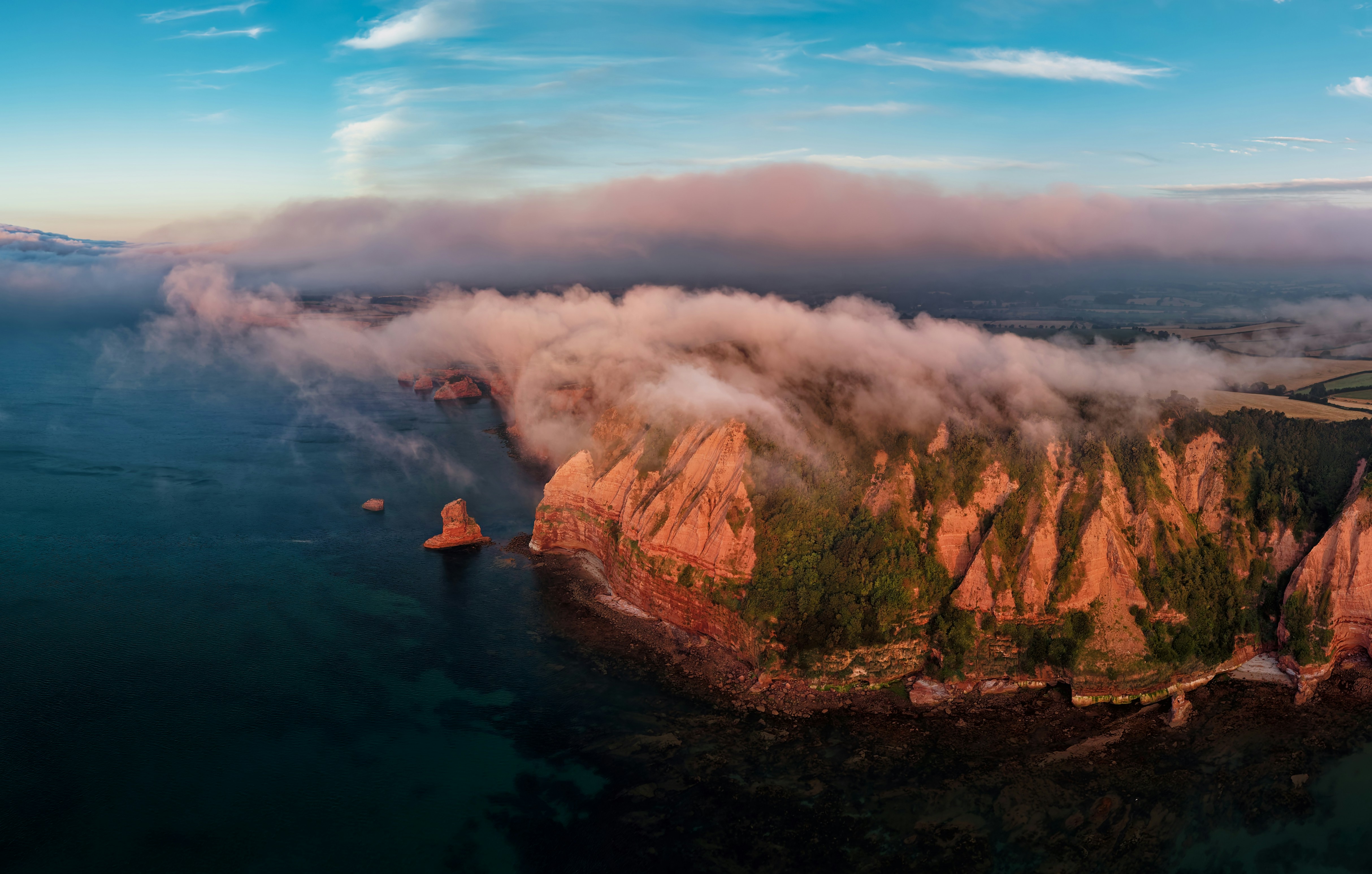 Sunlit limestone cliffs meet a calm sea with low-lying clouds drifting above; a panoramic coastal scene captured from an aerial vantage, highlighting rugged textures and soft atmospheric lighting.