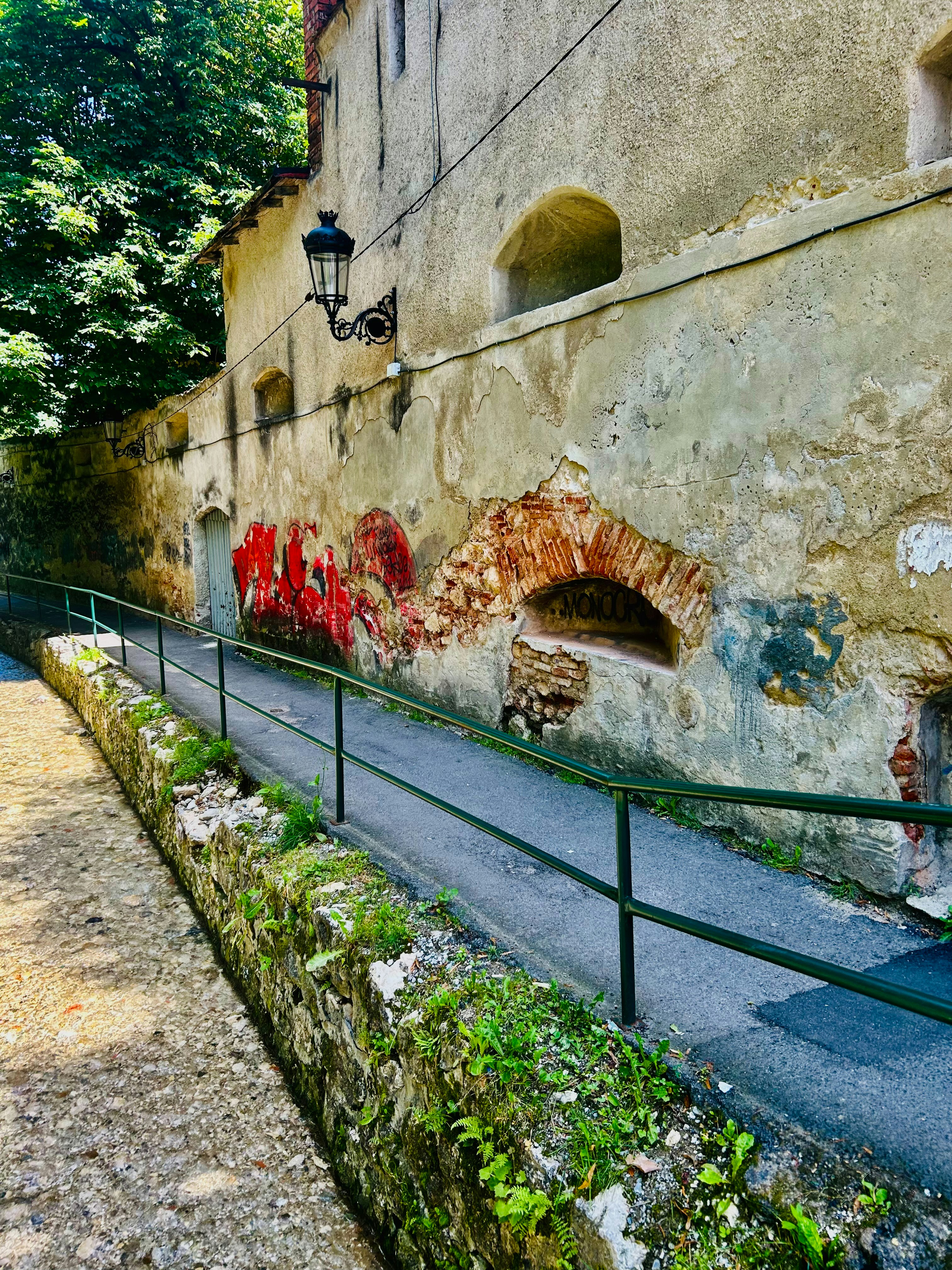 Weathered stone wall with a ramp and railing along a narrow pathway, showing remnants of red graffiti and a brick arch, captured beside a mossy ledge.