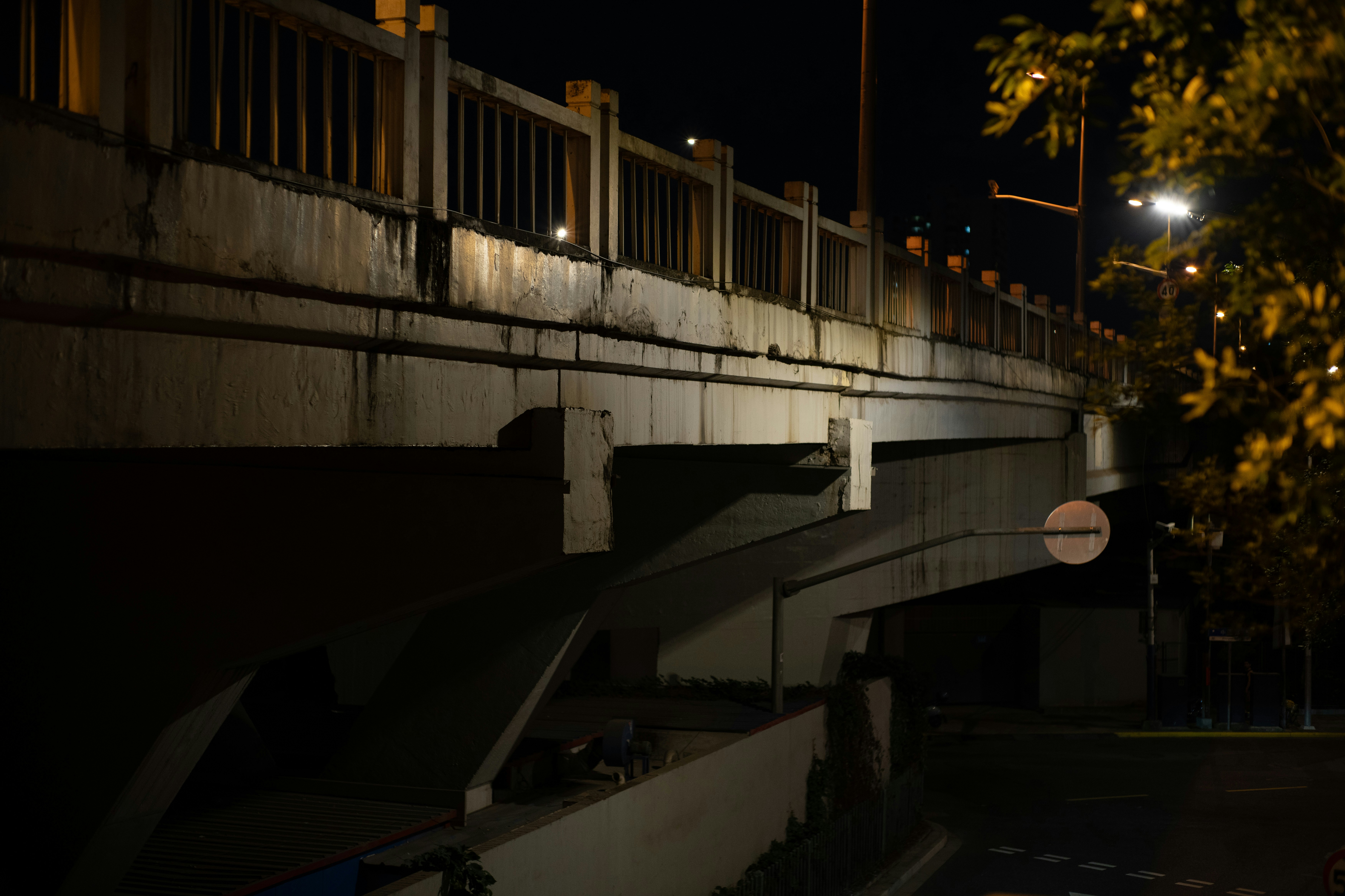 Dimly lit urban scene under a bridge with stairway shadows and distant streetlight; indicates a quiet nighttime passage and architectural concrete forms.