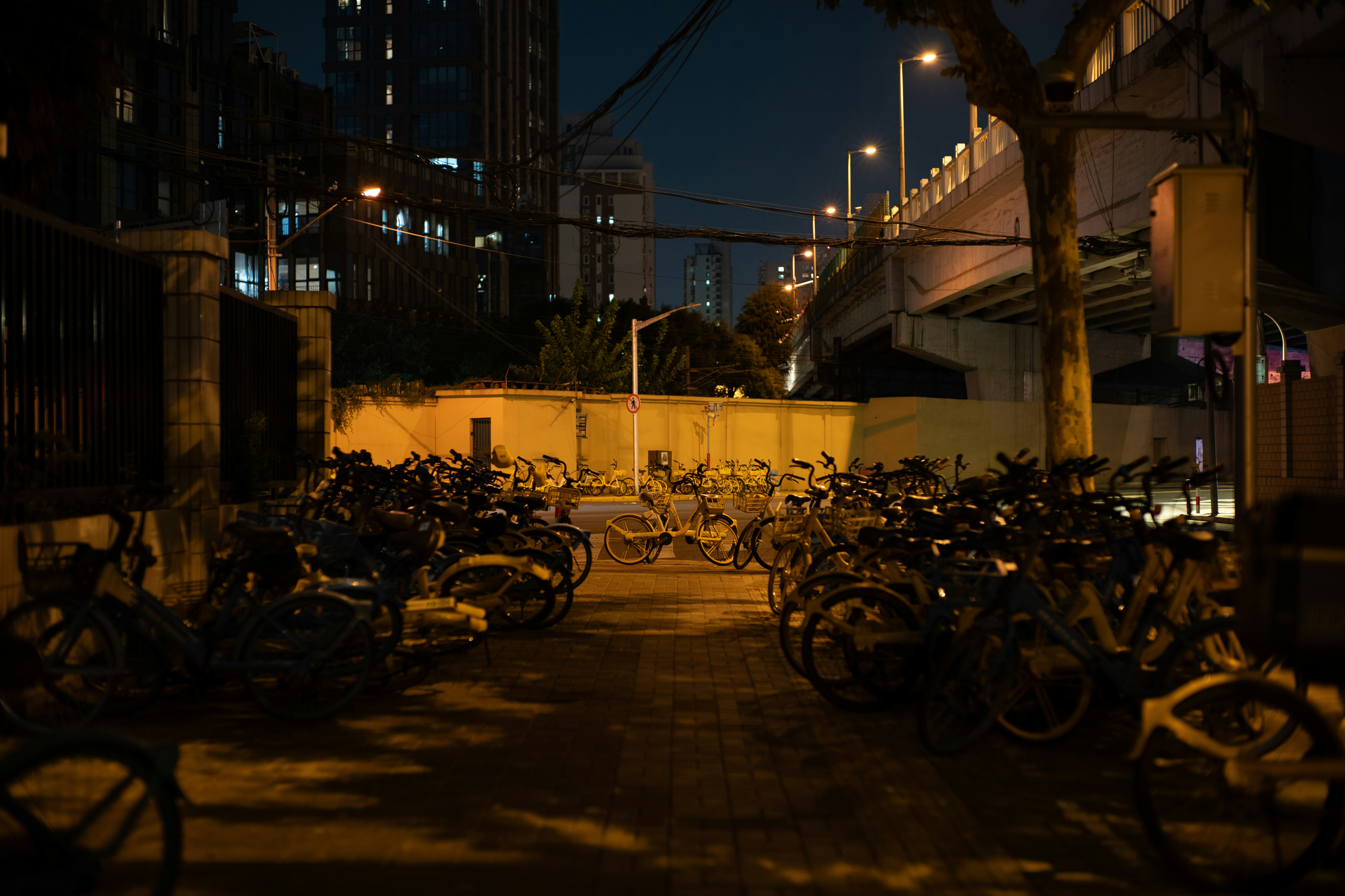 Dimly lit urban courtyard with bicycles lined along the path, illuminated by warm streetlights near an elevated roadway at night.
