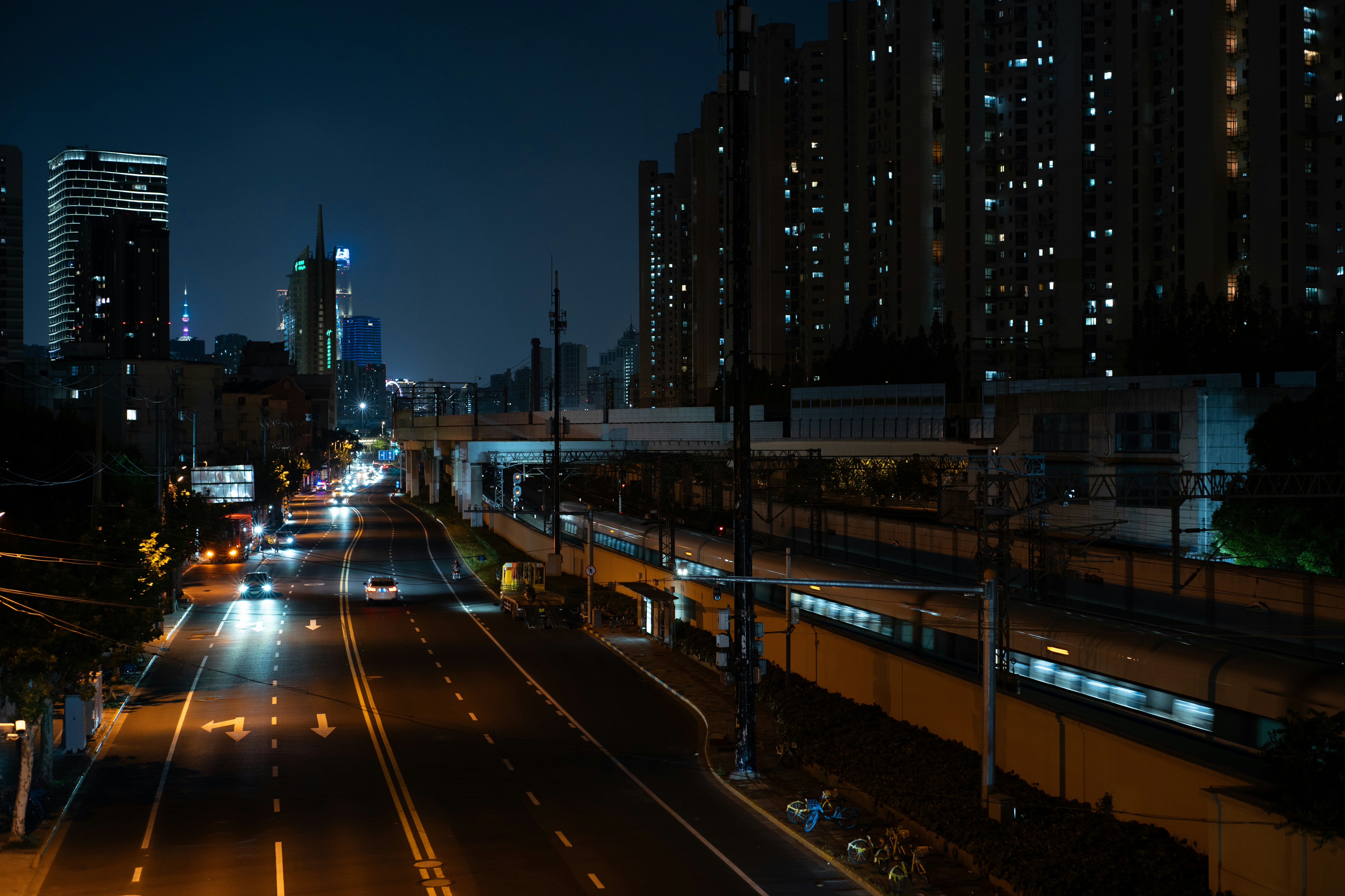 Nighttime city street with illuminated storefronts and tall apartment blocks along a straight avenue; distant traffic glows with car headlights and a moving train line on the right. Aerial perspective emphasizes urban rhythm and depth.
