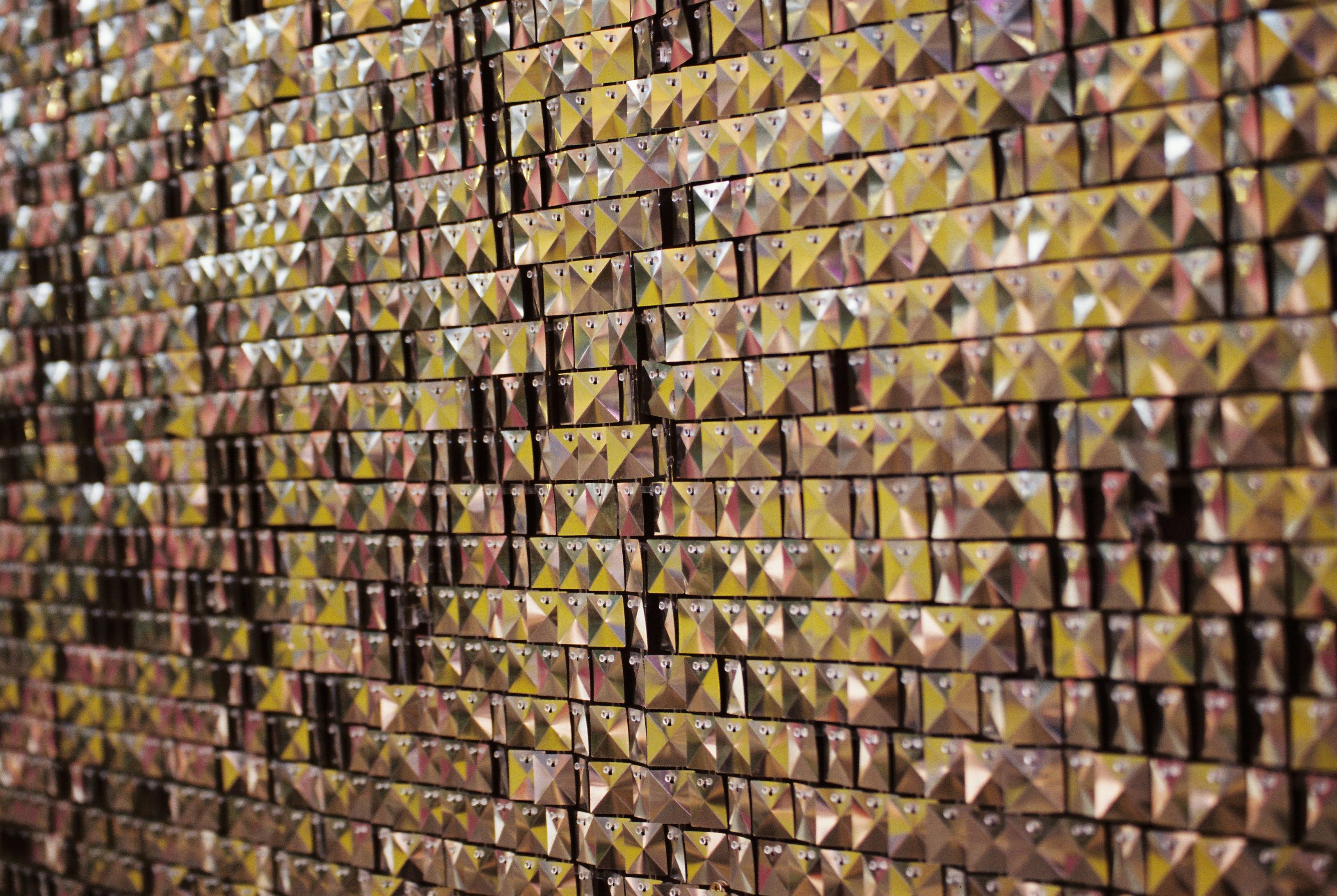Close-up of a textured wall composed of small, triangular brass tiles arranged in a repeating pattern, creating a metallic mosaic with warm amber tones.