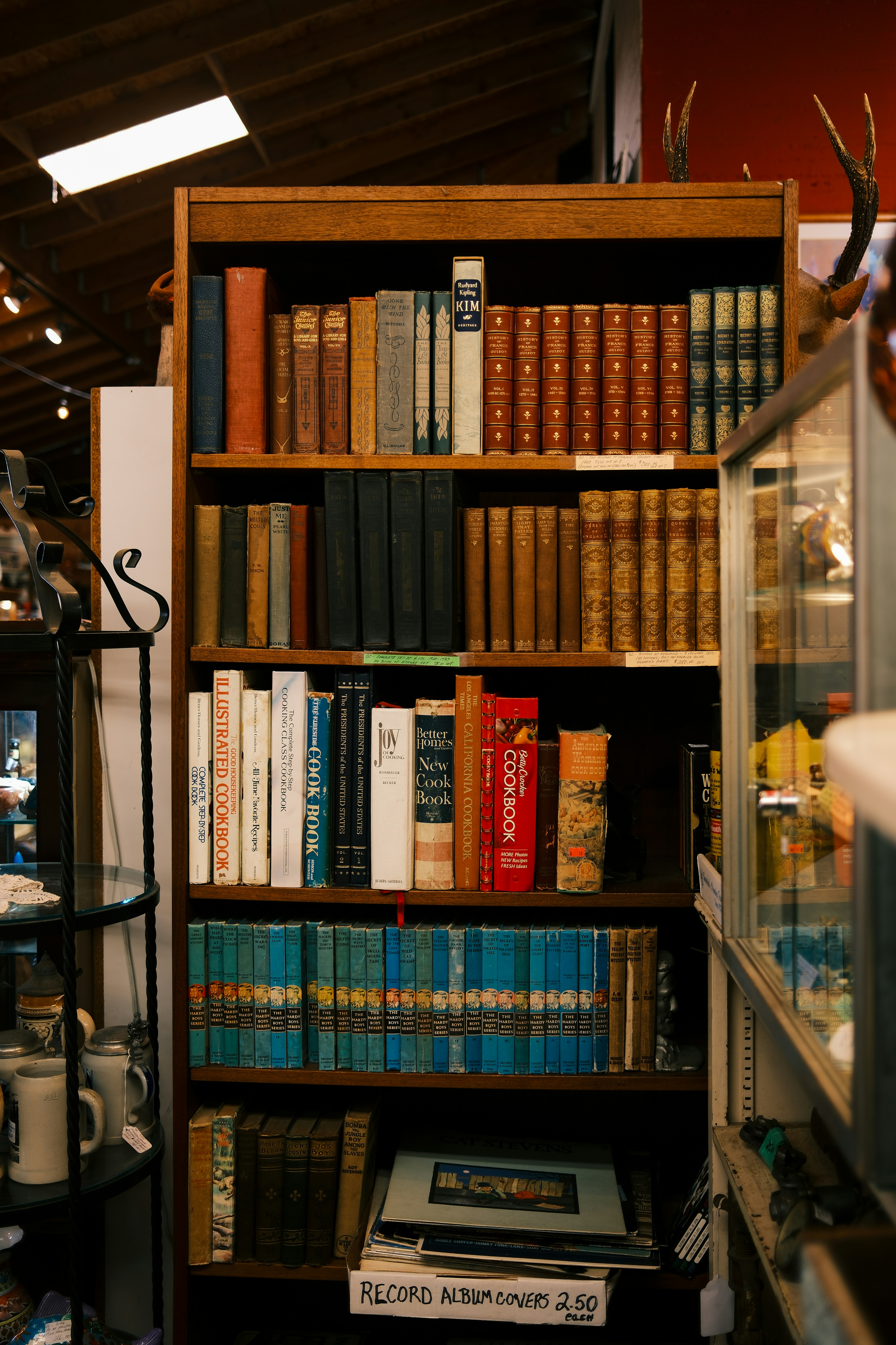 Tall wooden bookshelf filled with colorful old and new books in a cozy shop, with a glass display on the right and warm ambient lighting.