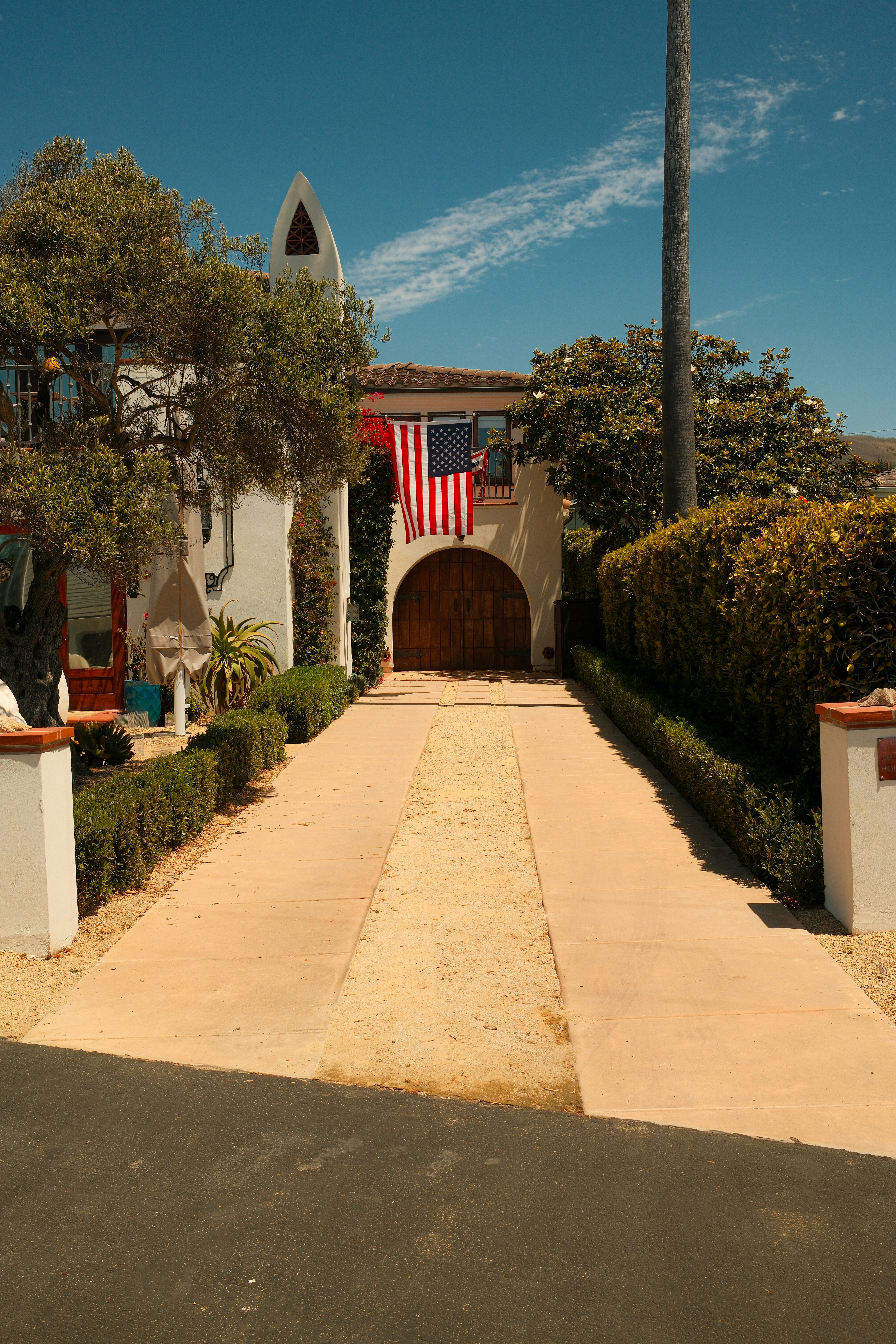Flag over a sunlit Spanish-style driveway leading to a arched wooden door