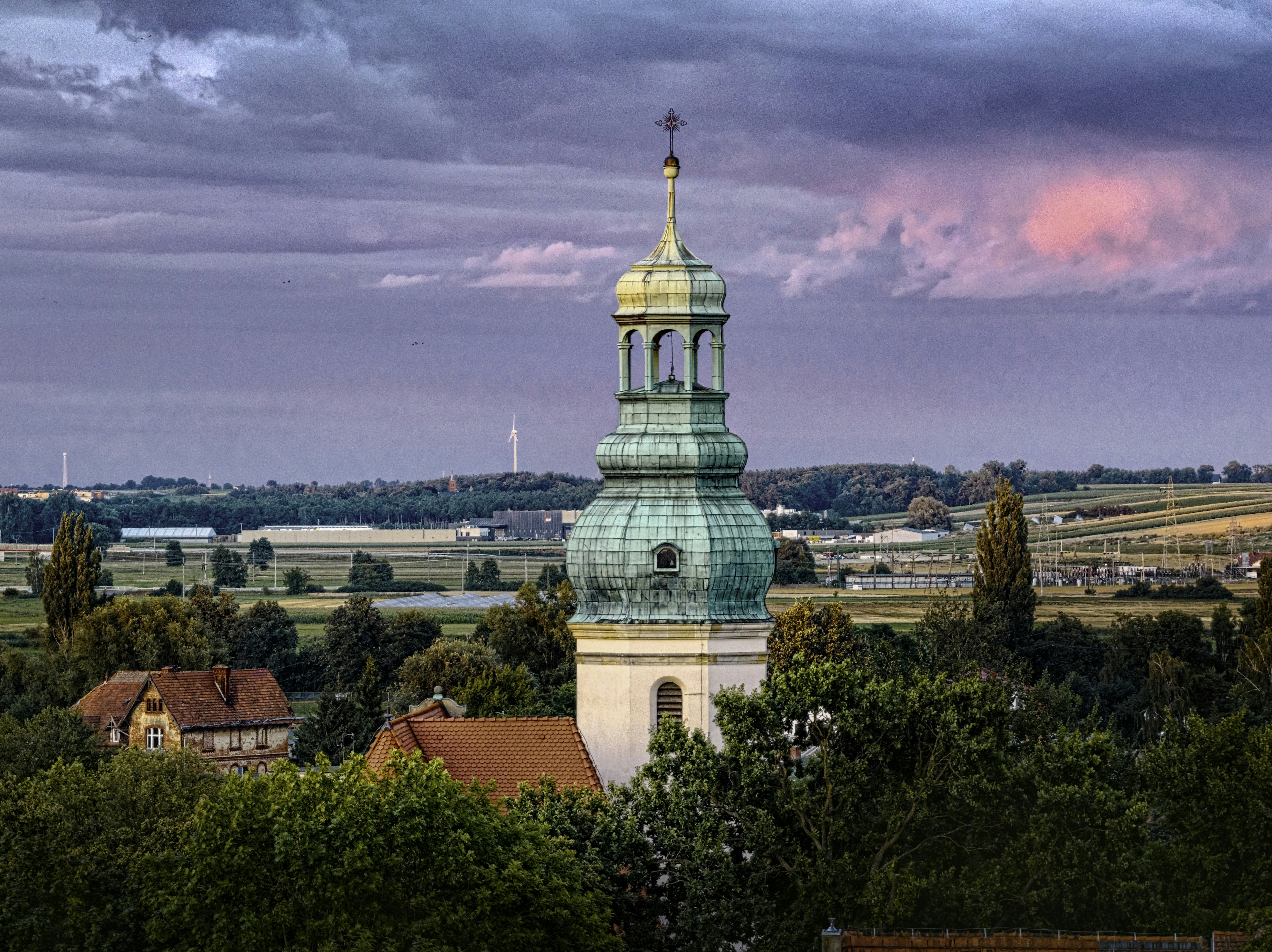 A green-domed church bell tower rises among trees and red-roofed houses with a distant countryside under a pink-tinged sky.
