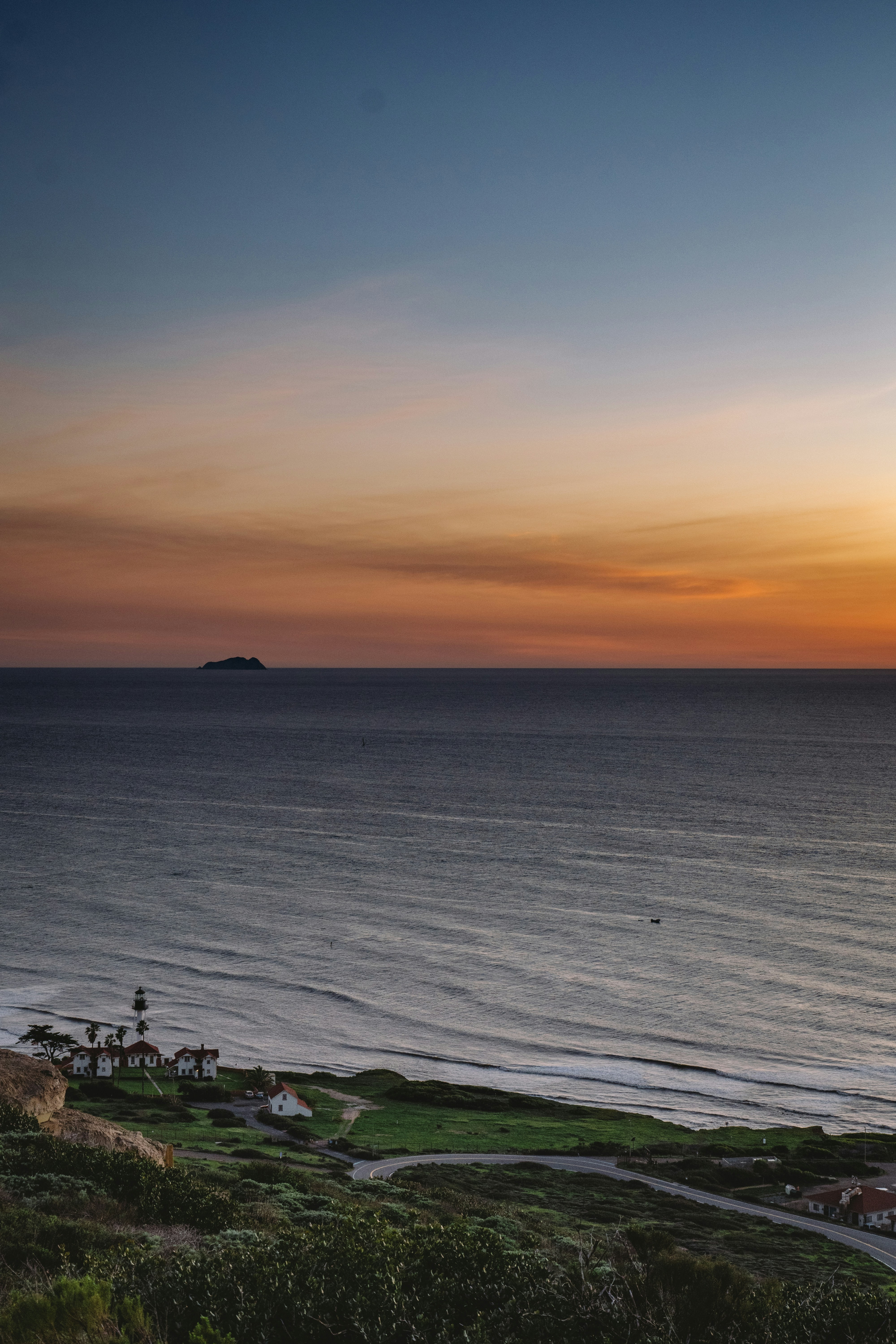 A coastal landscape at sunset with a small village in the foreground and calm sea stretching to a distant island, the sky glowing orange and blue above. This is a photographic capture of a serene coastline.