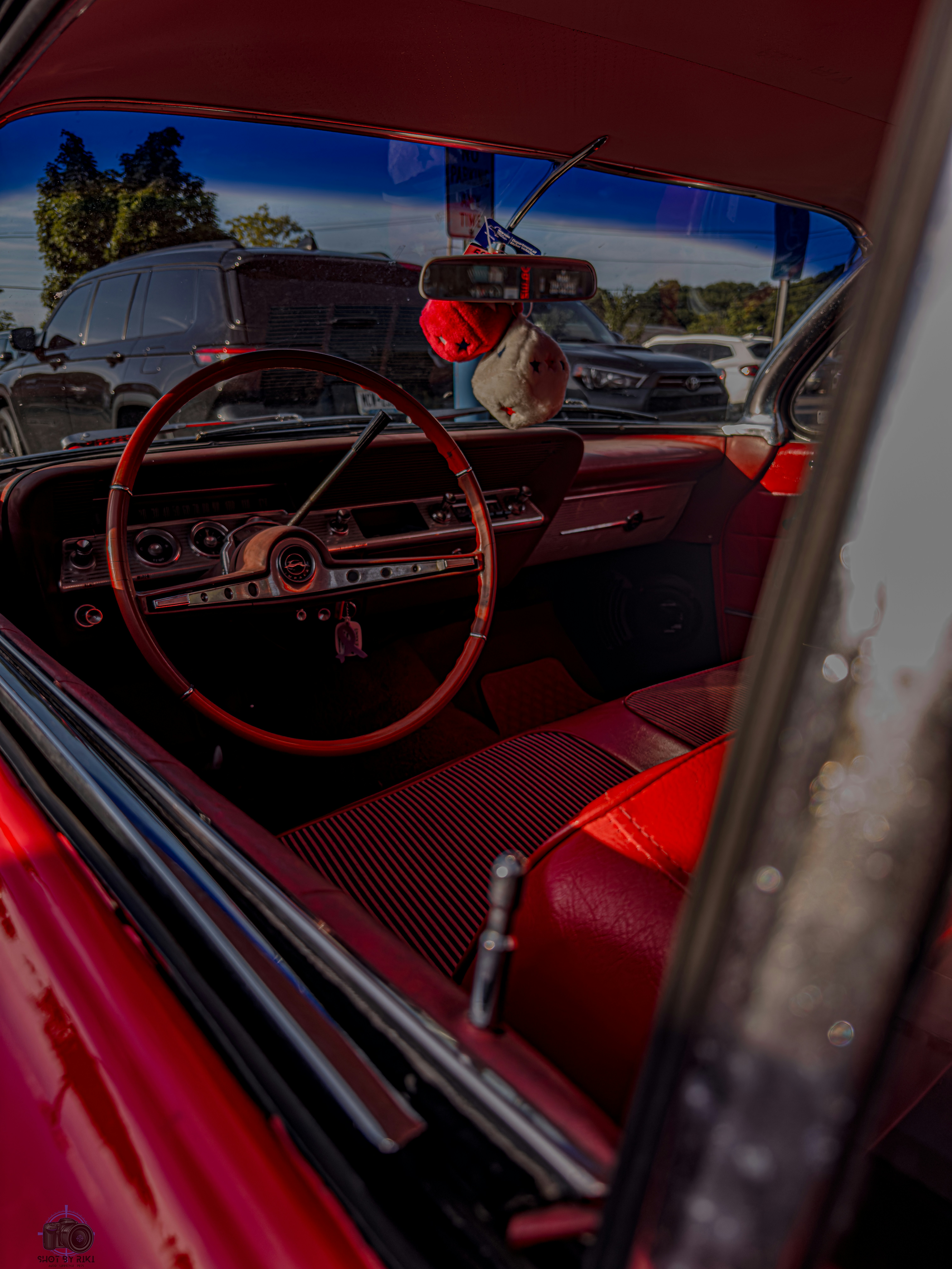Red upholstery and steering wheel fill a vintage car interior with sunlit details; hanging fuzzy ornaments add a personal touch to the scene.