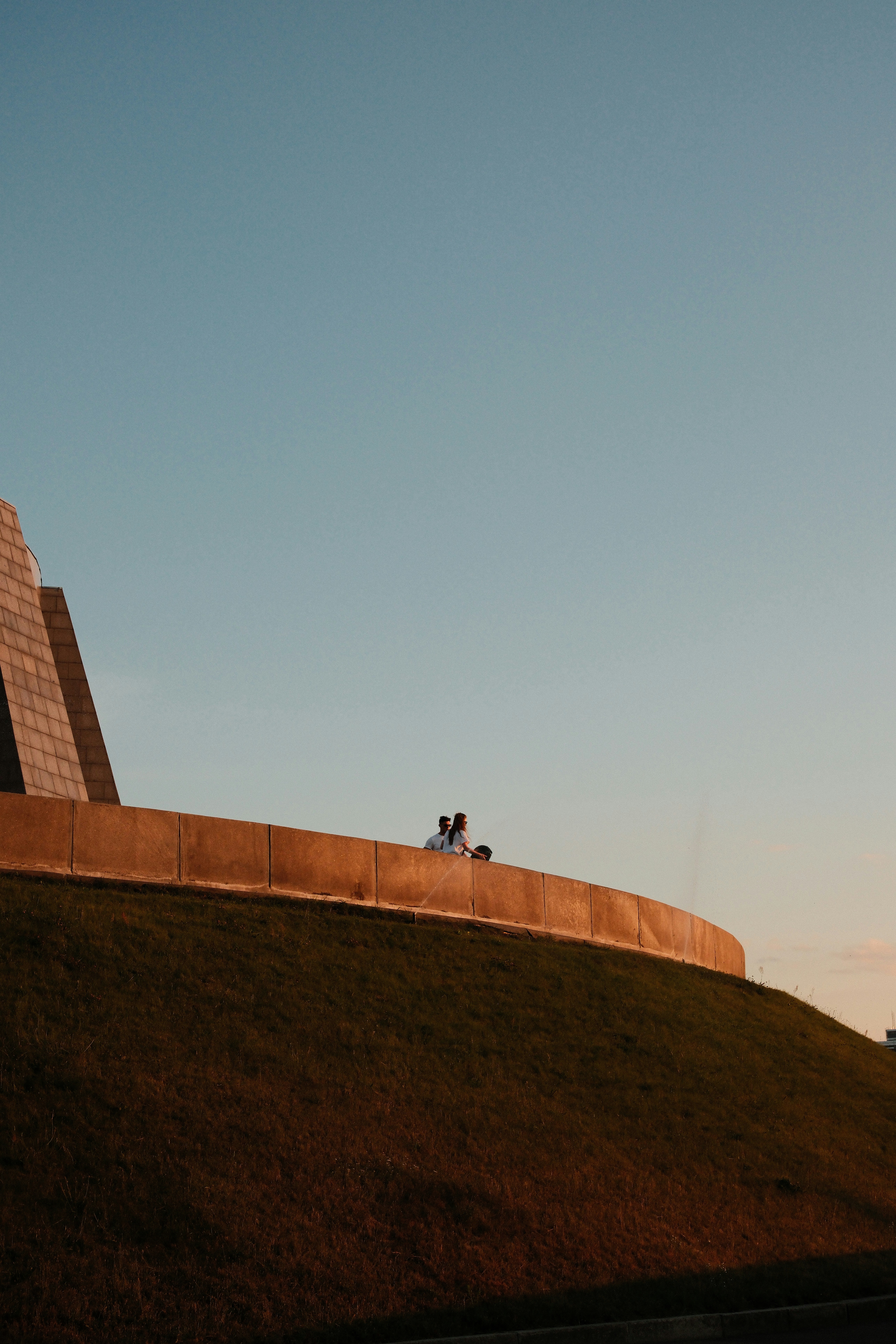 Two figures lean on a curved barrier at the edge of a grassy slope under a clear sky, captured at golden hour with warm light highlighting their silhouette.
