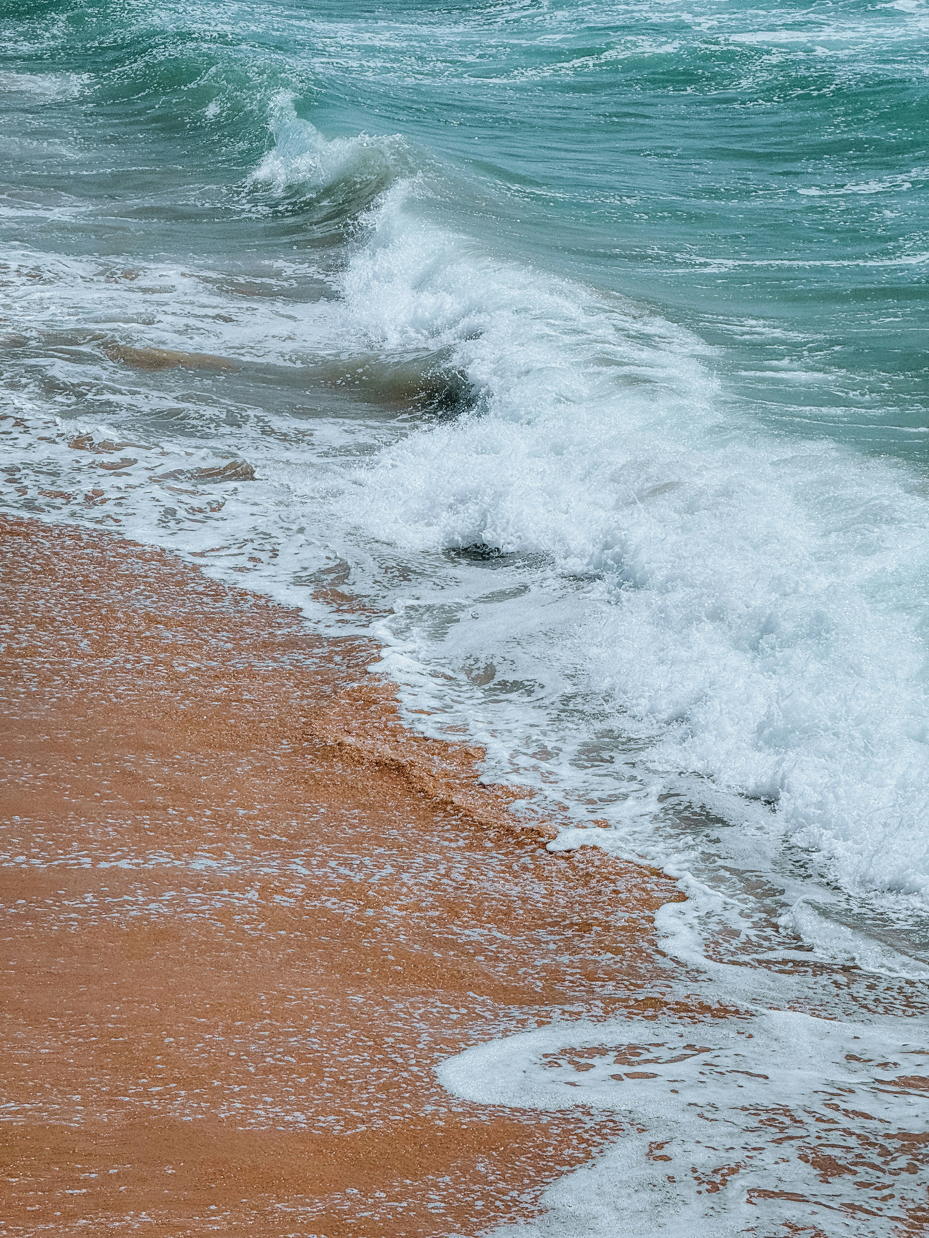 Waves crash along a sunlit sandy beach, with turquoise water meeting the orange-brown shore in frothy spray. A calm seascape emphasizing the shoreline transition.