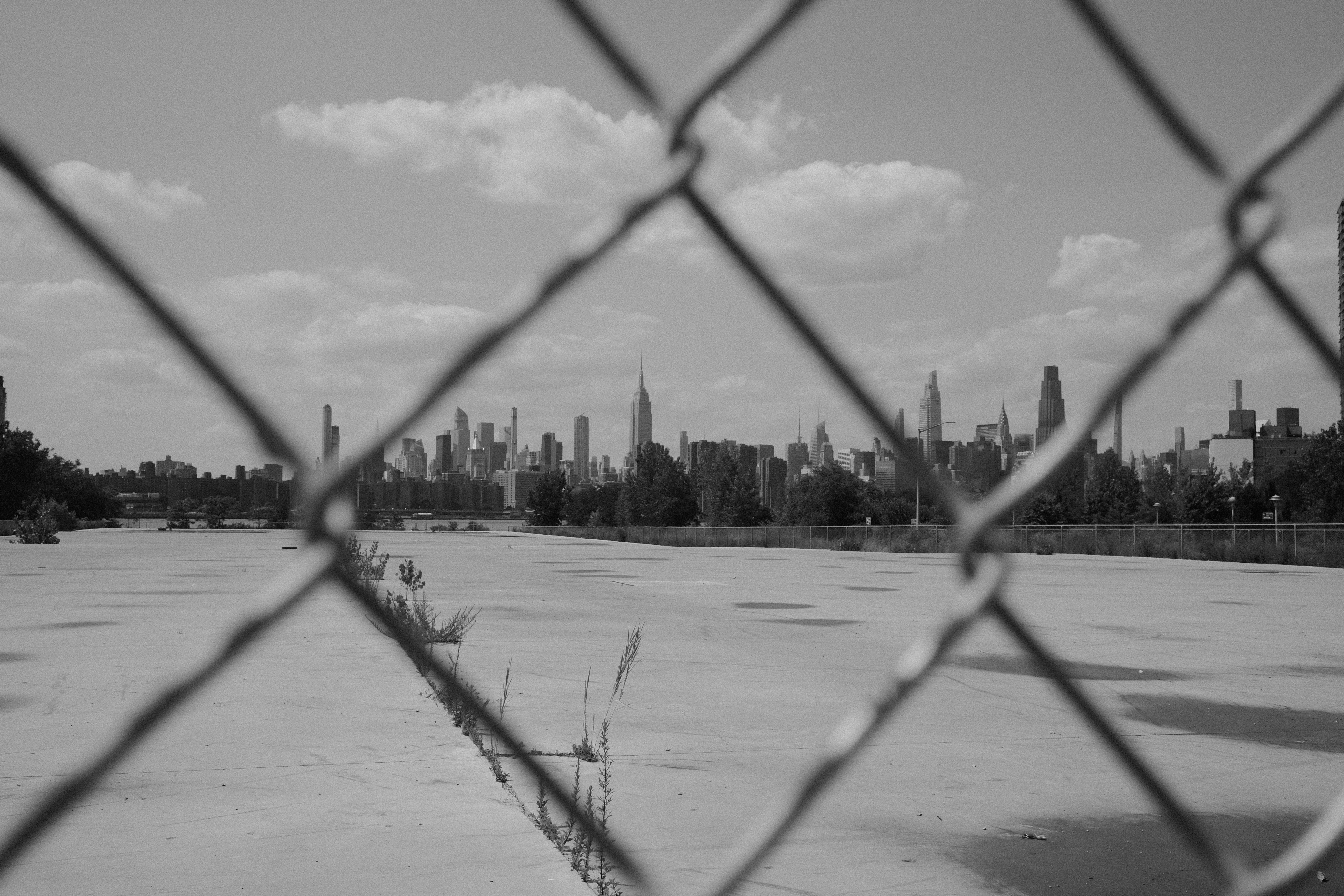 A black-and-white urban skyline viewed through a chain-link fence, with a frozen, reflective foreground and a distant cityscape on the horizon.