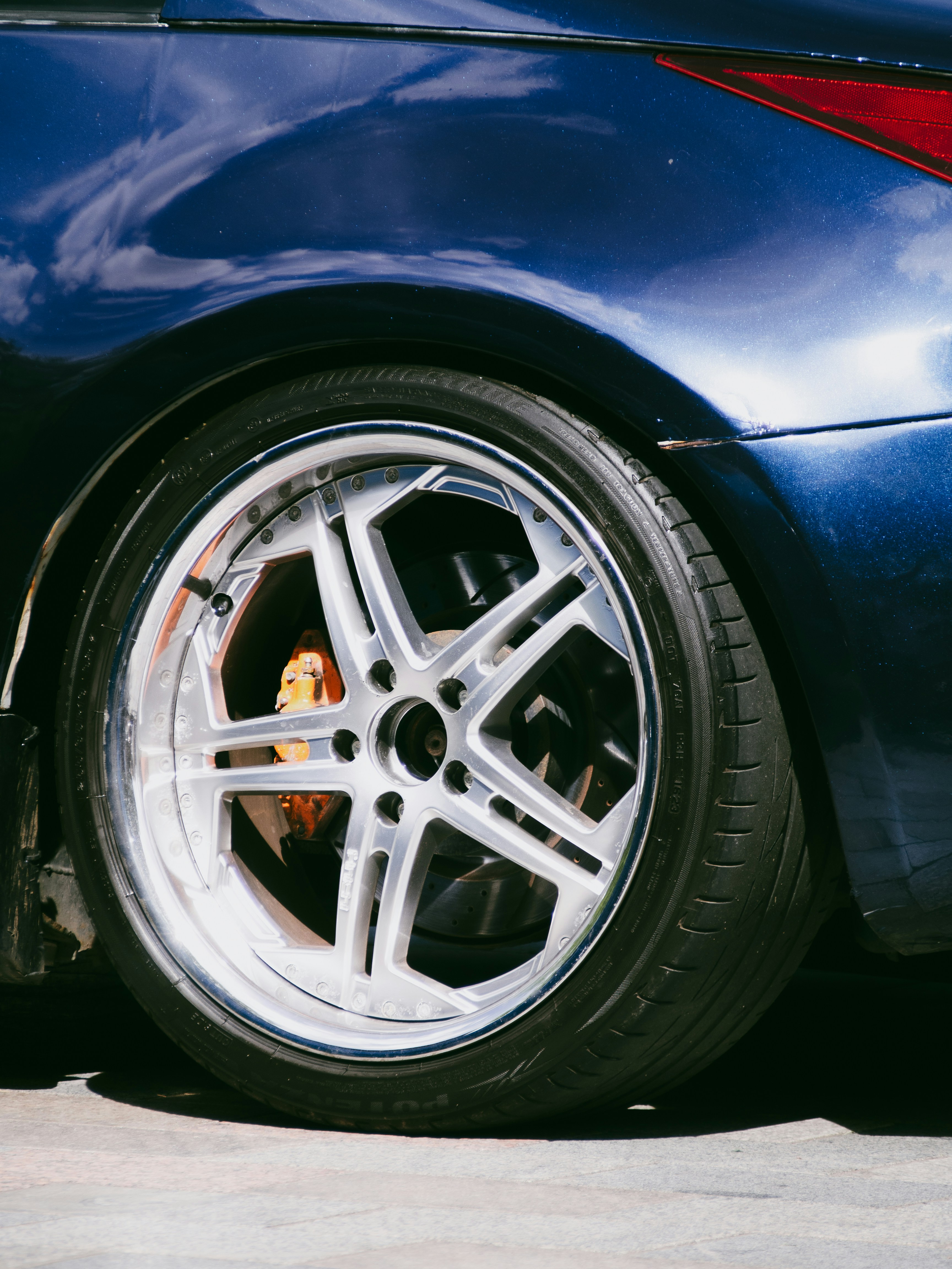 Close-up of a metallic alloy wheel and brake caliper inside a blue car fender, highlighting polished spokes and tire tread; non-mobile composition, with visible reflection on the body and pavement texture. This is a photo of a real car part.