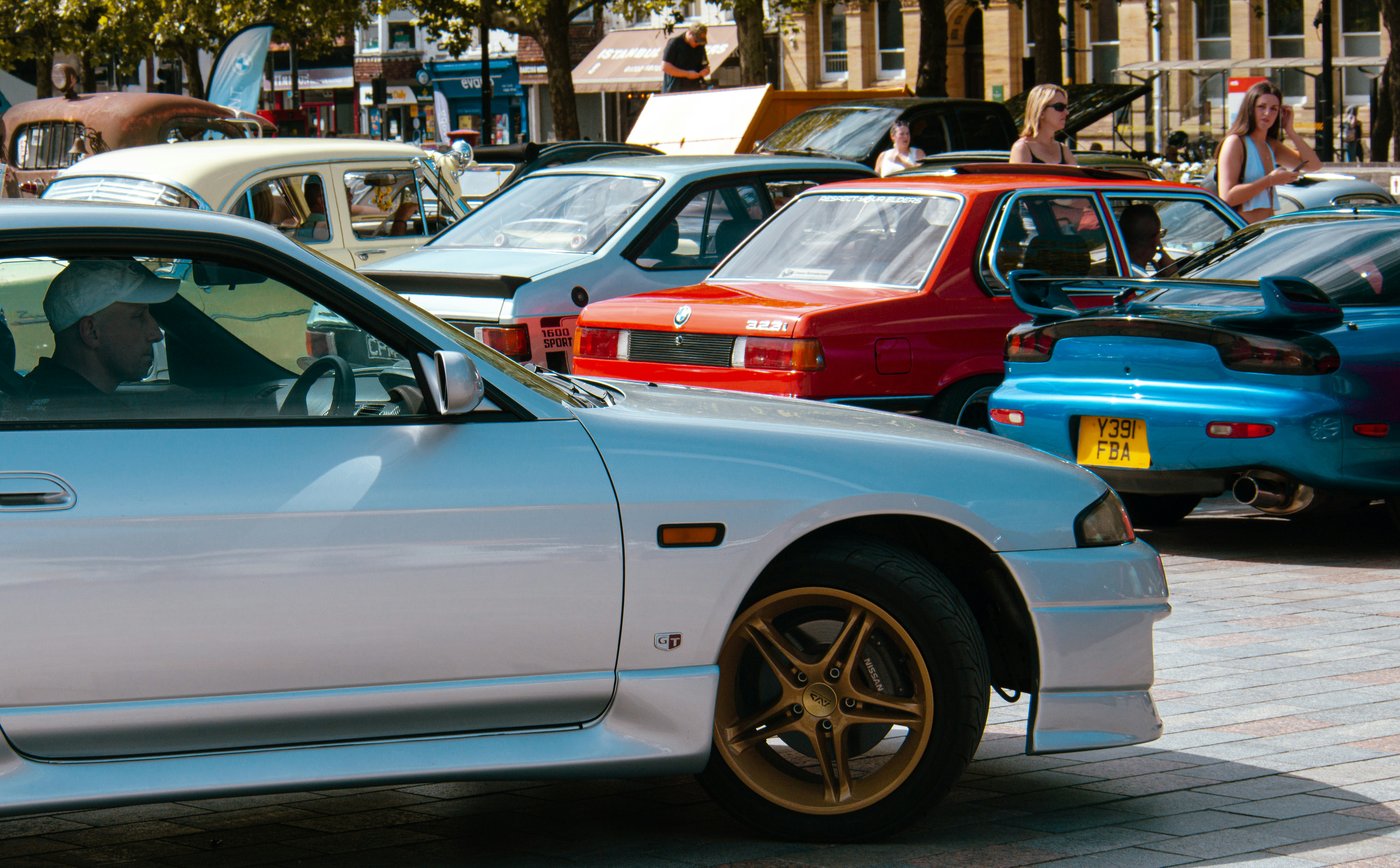 Bright blue sports coupe front and center among a row of colorful classic cars in a sunny street display. People and storefronts frame the background.