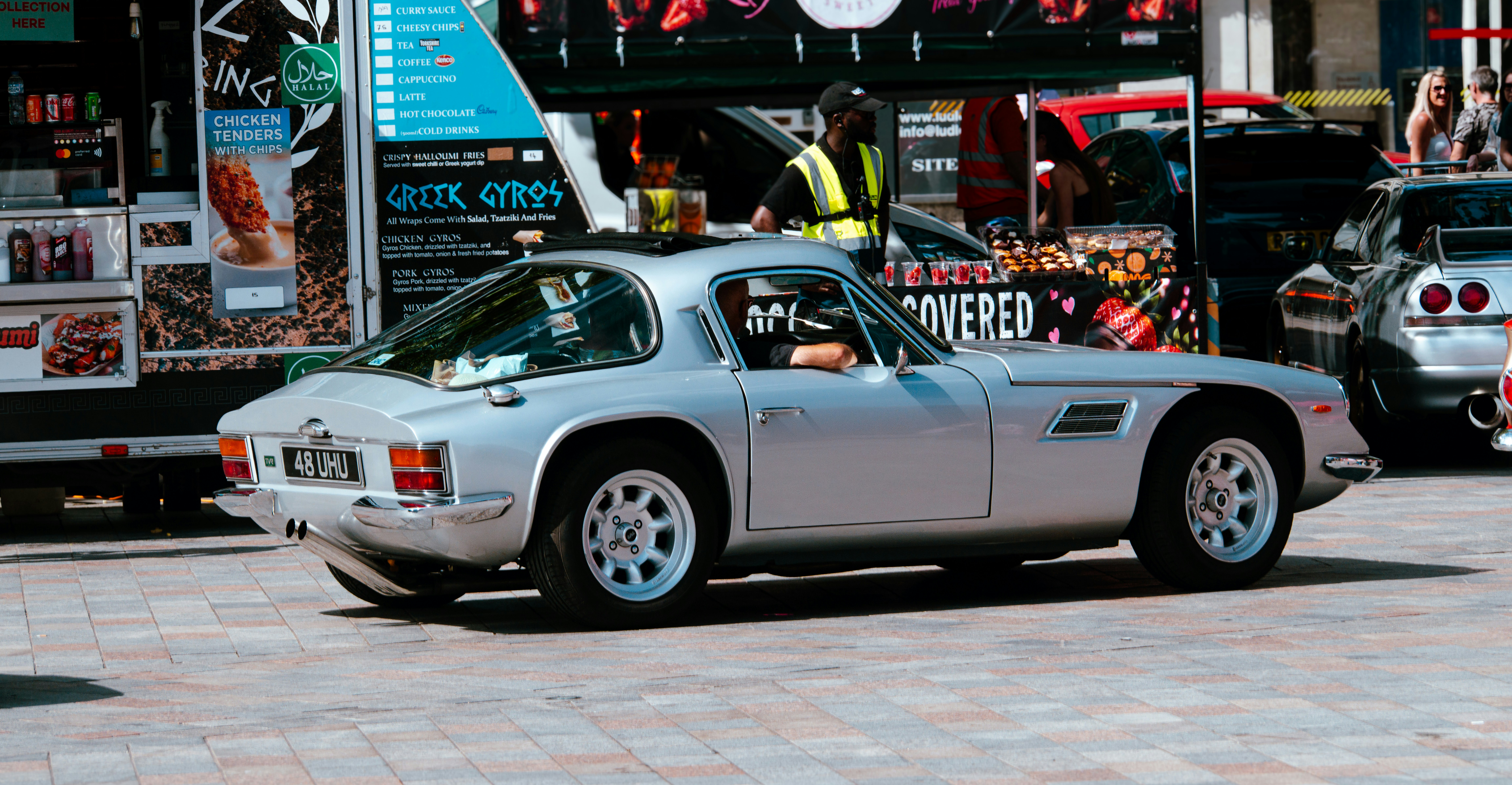 A silver vintage Porsche sits in a busy urban square with food stalls and people in the background, showcasing a contrast between classic design and modern commerce.
