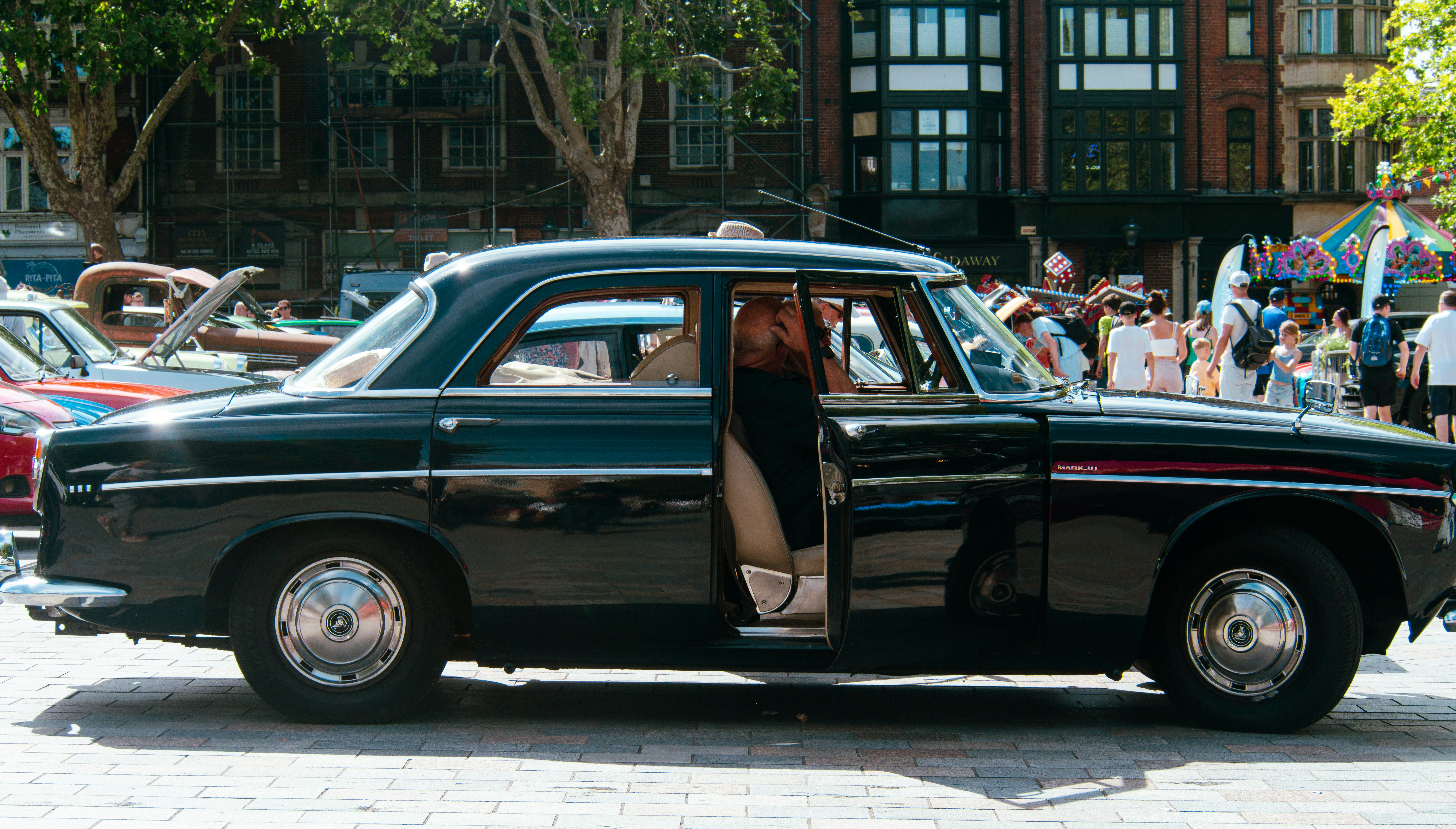 A vintage black car with an open rear door sits on a sunny street, crowd and stalls in the background; a retro automobile at a public event.