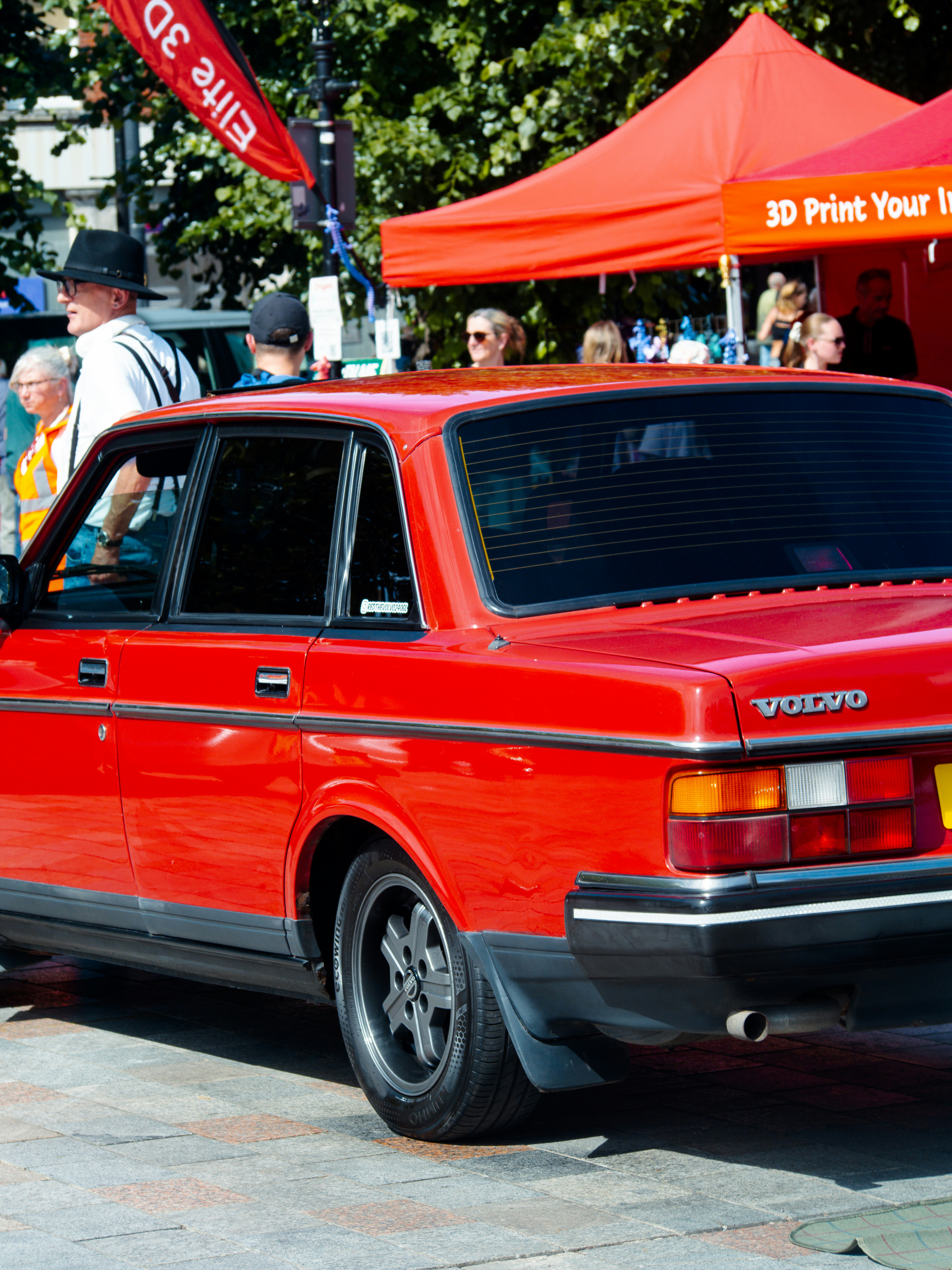 Bright red Volvo sedan at an outdoor event with tents and people in the background, showcasing a vintage car scene.