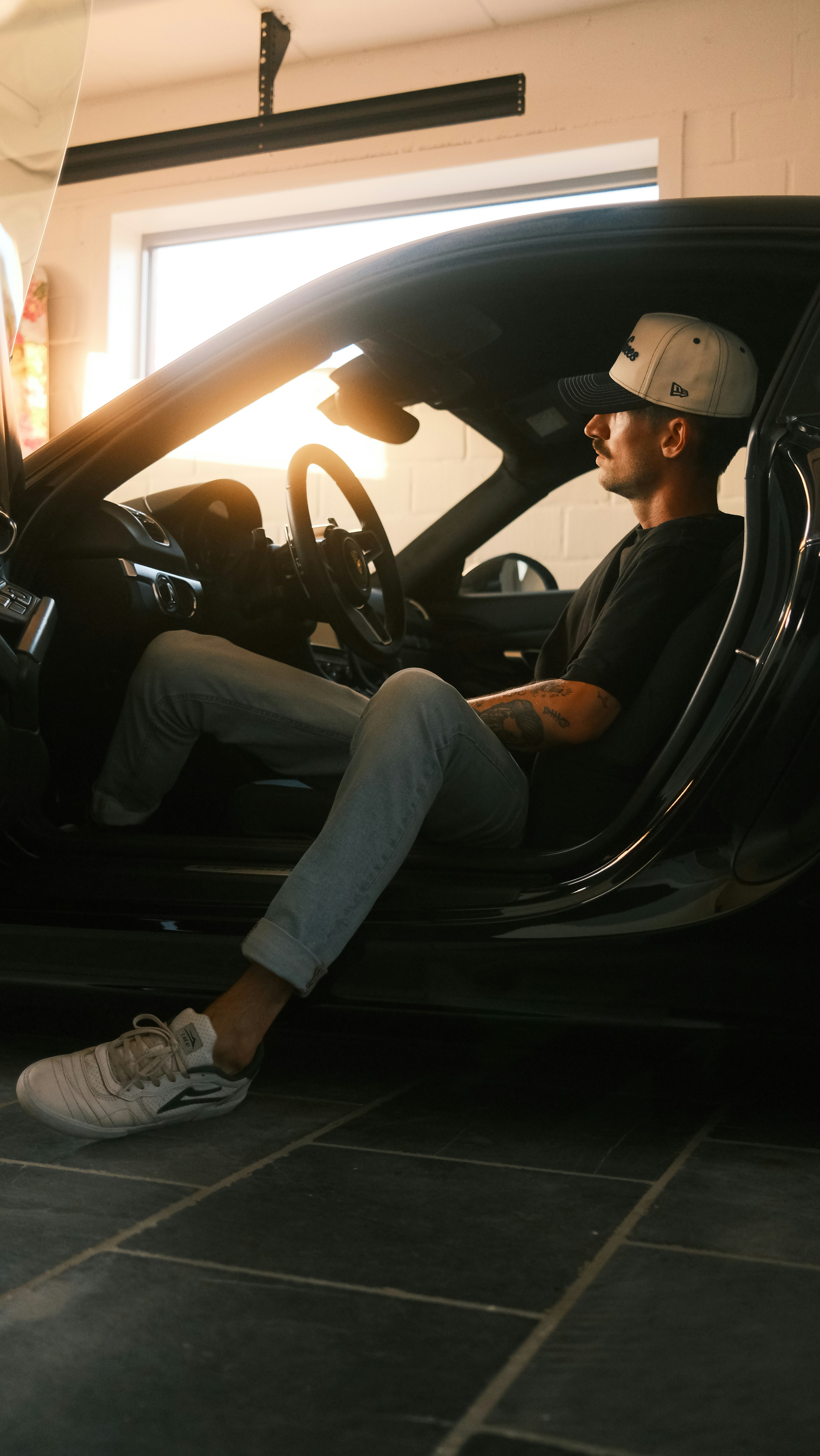 Man seated in a black sports car, wearing a white cap, jeans, and sneakers; sunlight from the window creates a warm glow inside the vehicle.