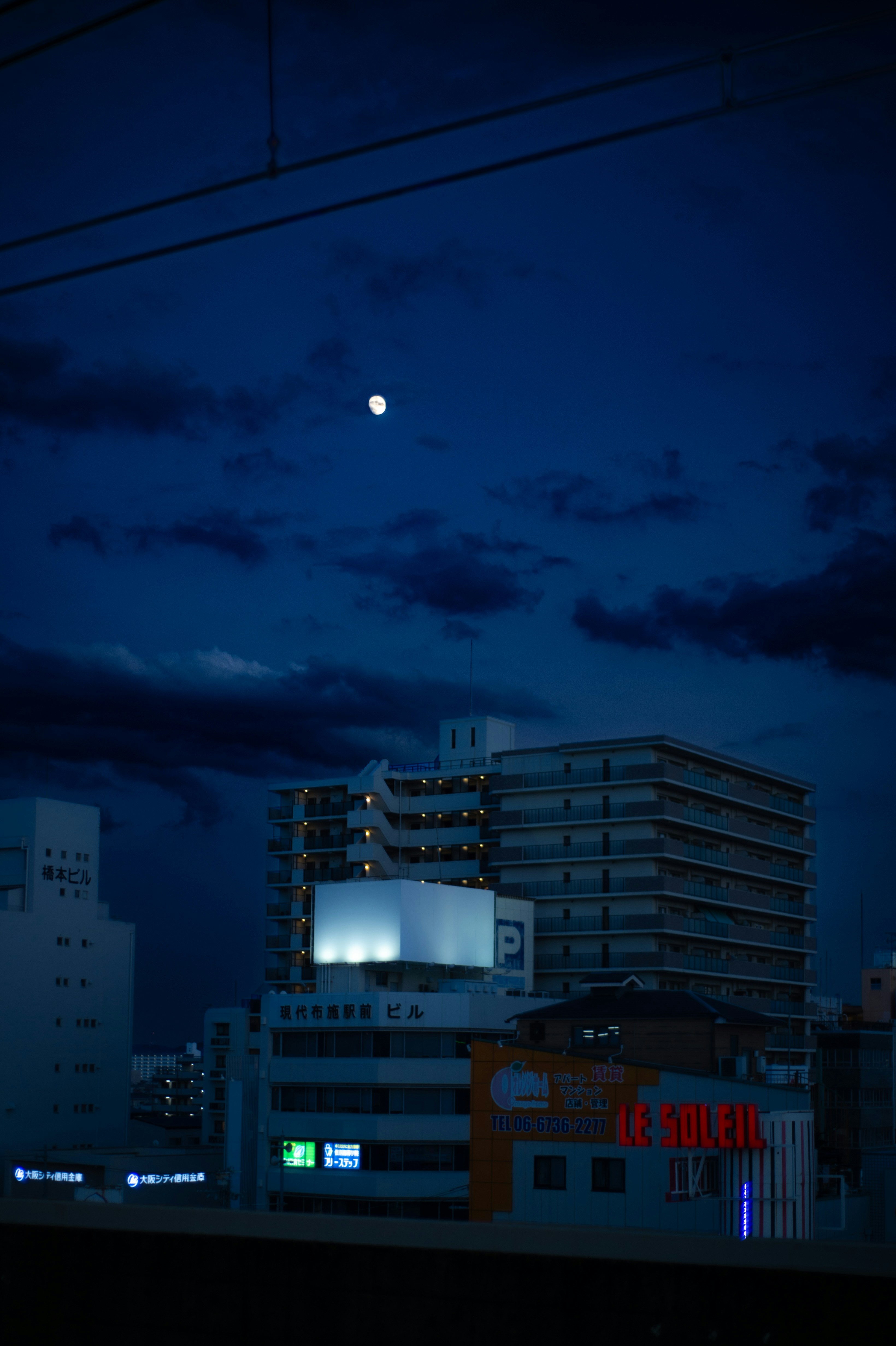 Moon hangs over a quiet cityscape of mid-rise buildings at dusk; the scene is bathed in cool blue tones with a few illuminated signs giving a hint of life below. This is a photographic capture featuring a skyline and subtle artificial lighting.