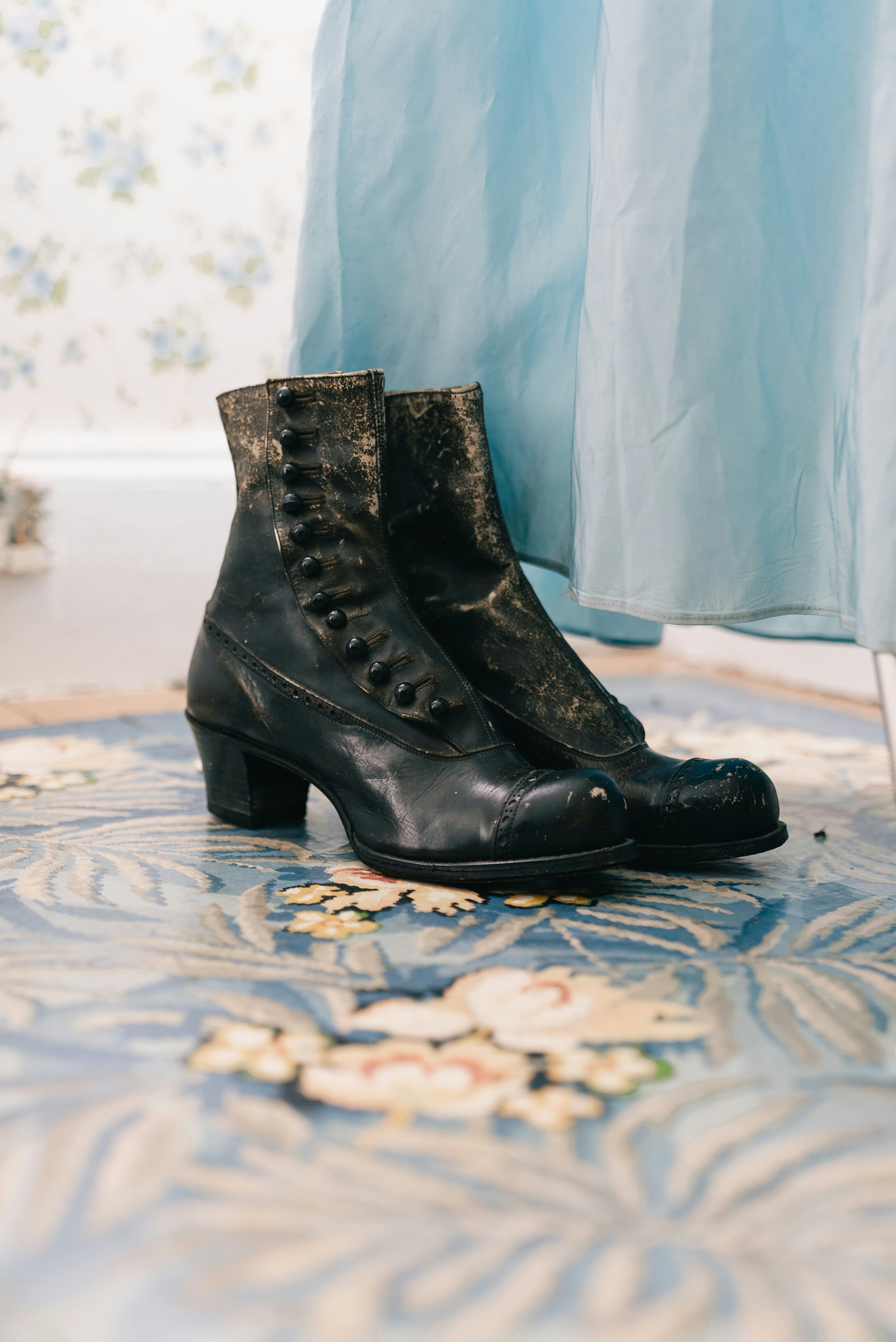 Pair of black ankle boots resting on a patterned rug under a pale blue curtain.