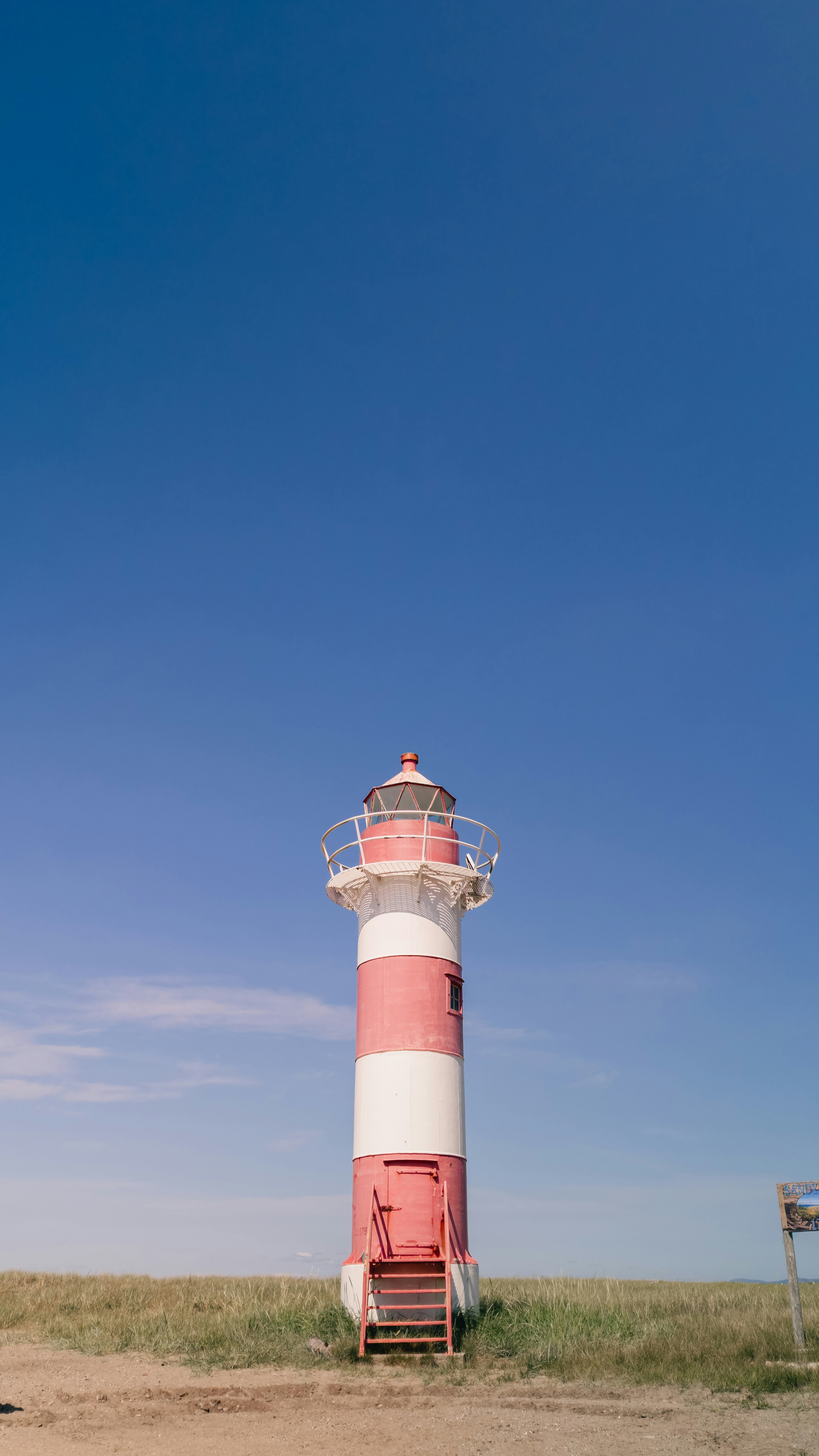 A striped lighthouse stands on a grassy shore beneath a vast blue sky, with a small stairs leading up to its door and a distant sign to the right.
