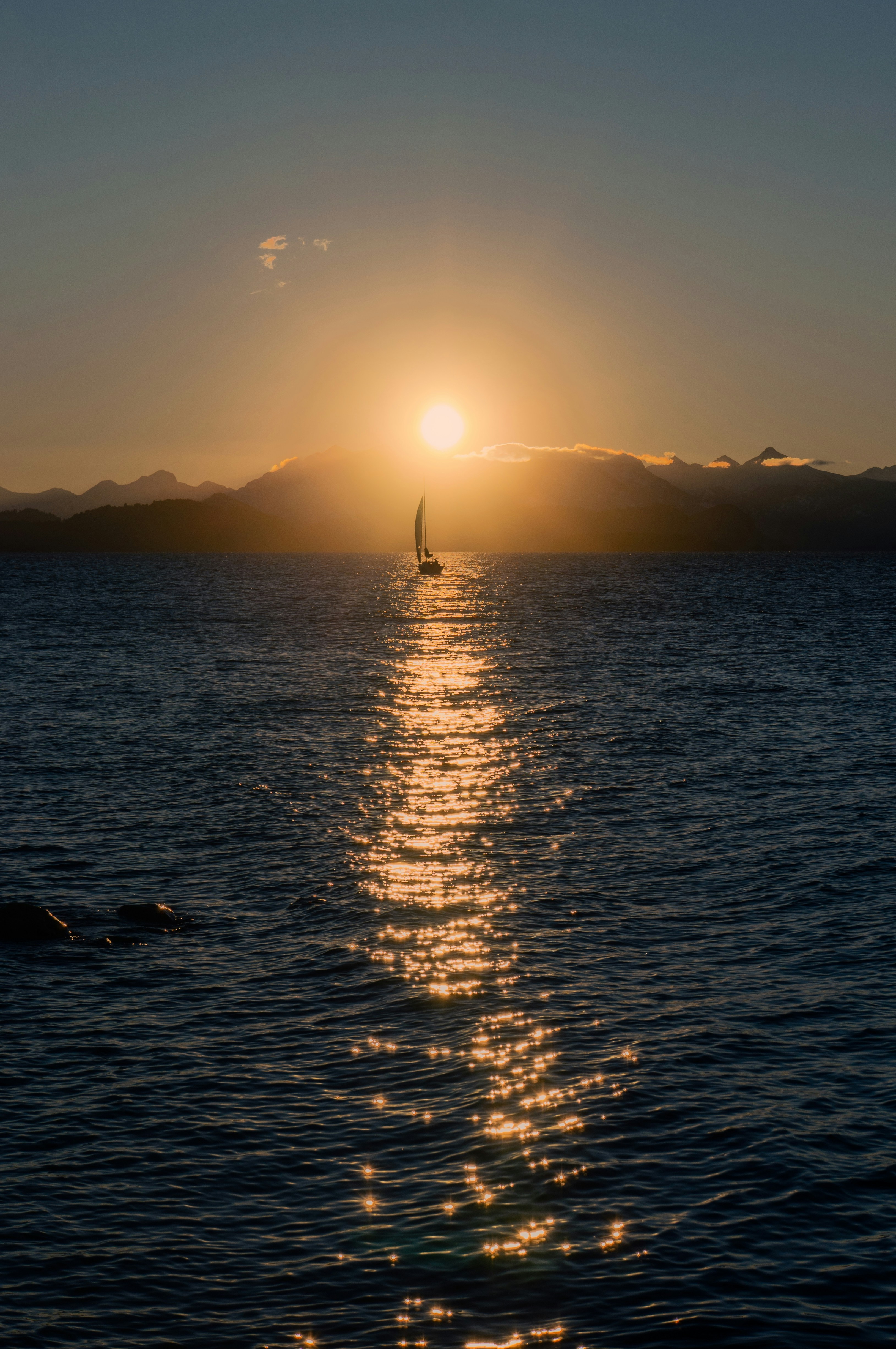 Golden sunset over calm water with a lone sailboat near the horizon; the sun creates a bright path on the sea. A serene seascape, captured as a natural landscape photograph.