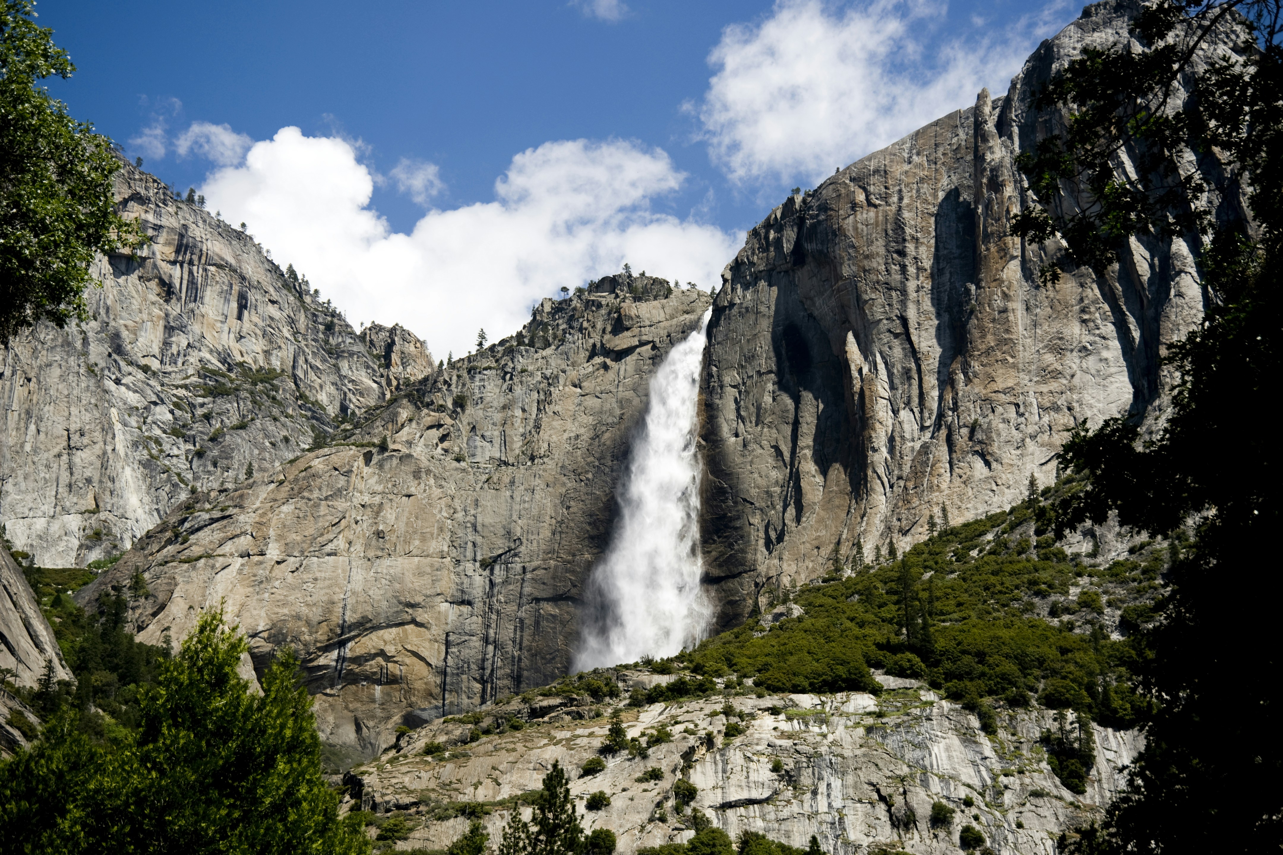 Sturdy canyon scene featuring a tall waterfall cascading between rugged granite cliffs; pine trees dot the lower slopes, under a bright blue sky with scattered clouds.